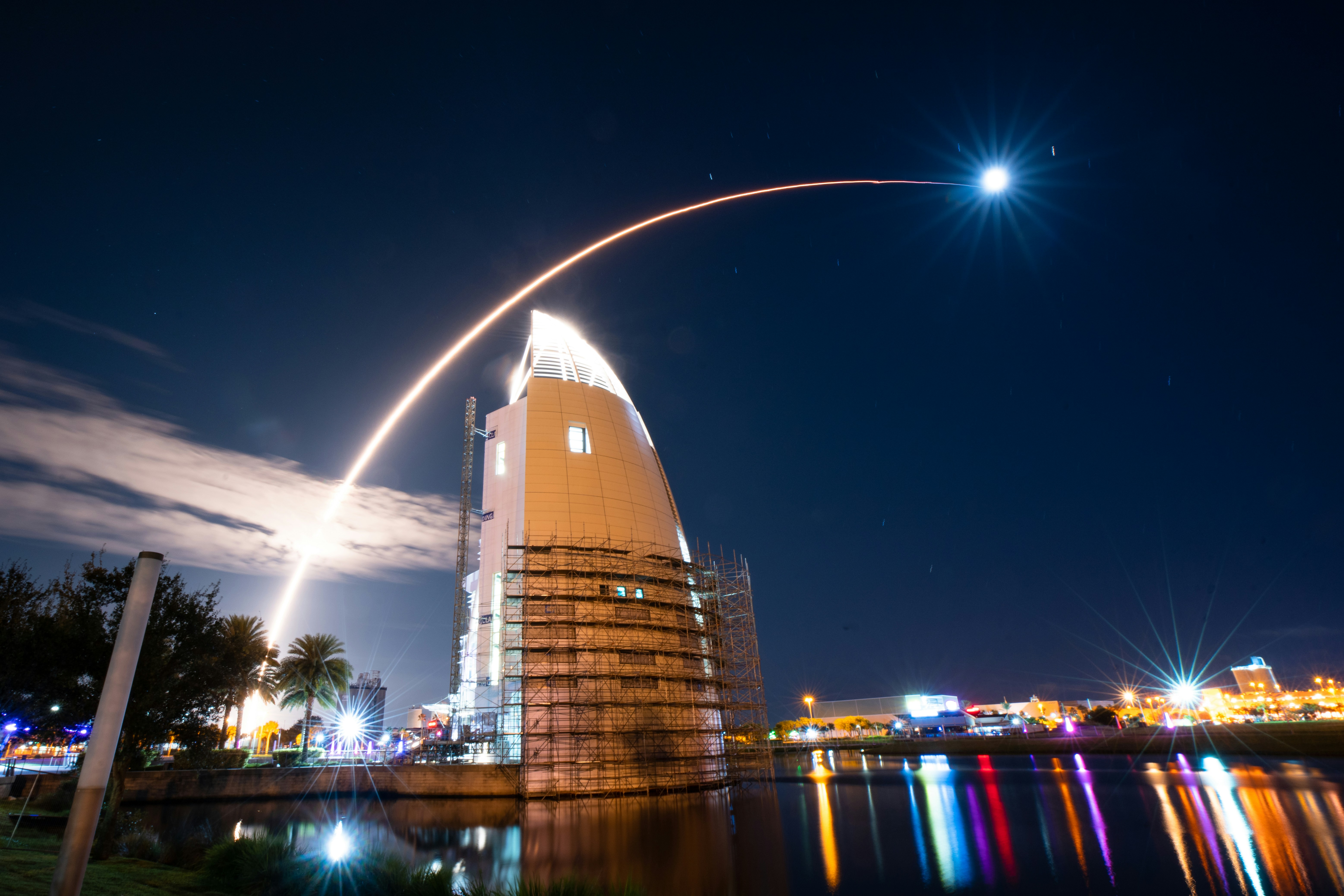 Rocket trajectory illuminating the night sky above a partially constructed building, reflecting in the water below.