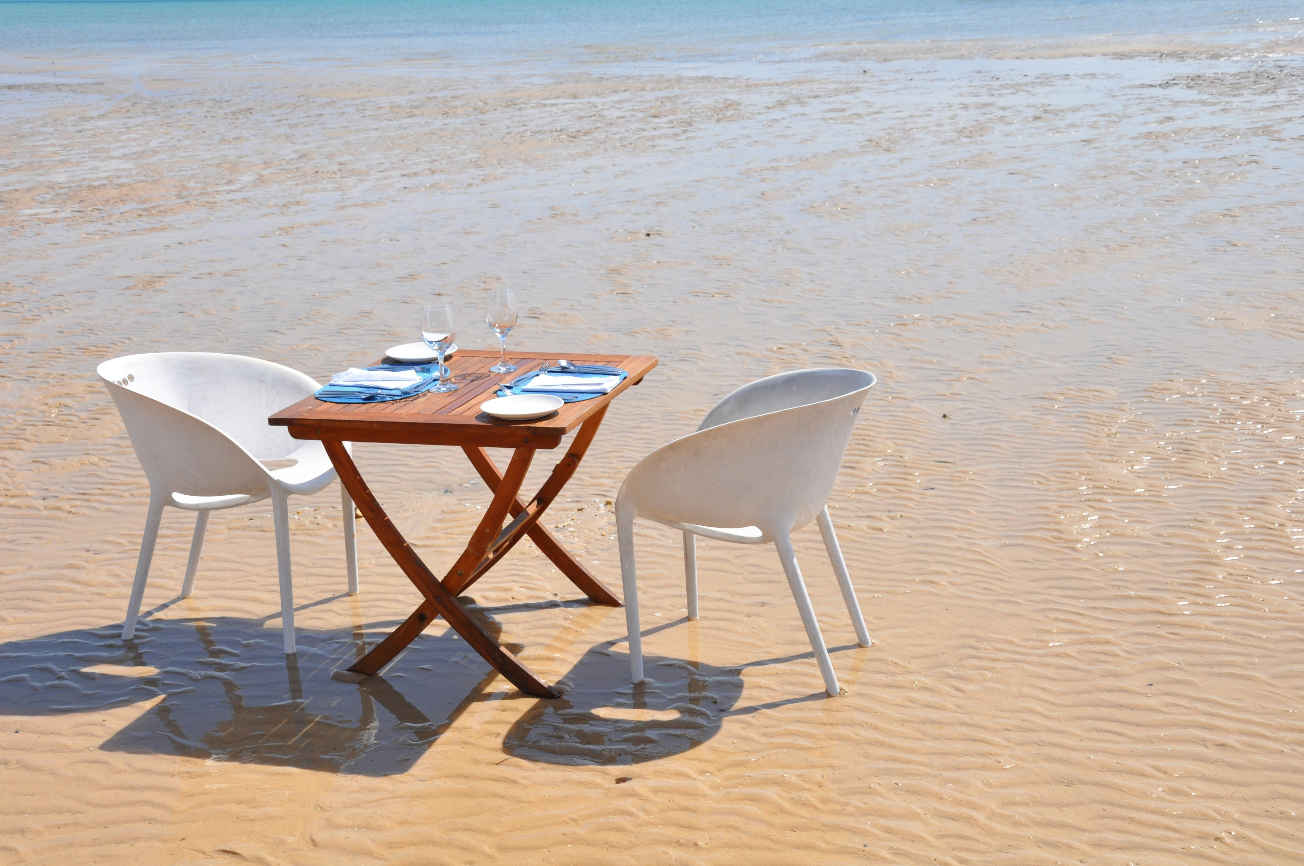 Two white chairs and a wooden table set for breakfast on a sunlit beach.