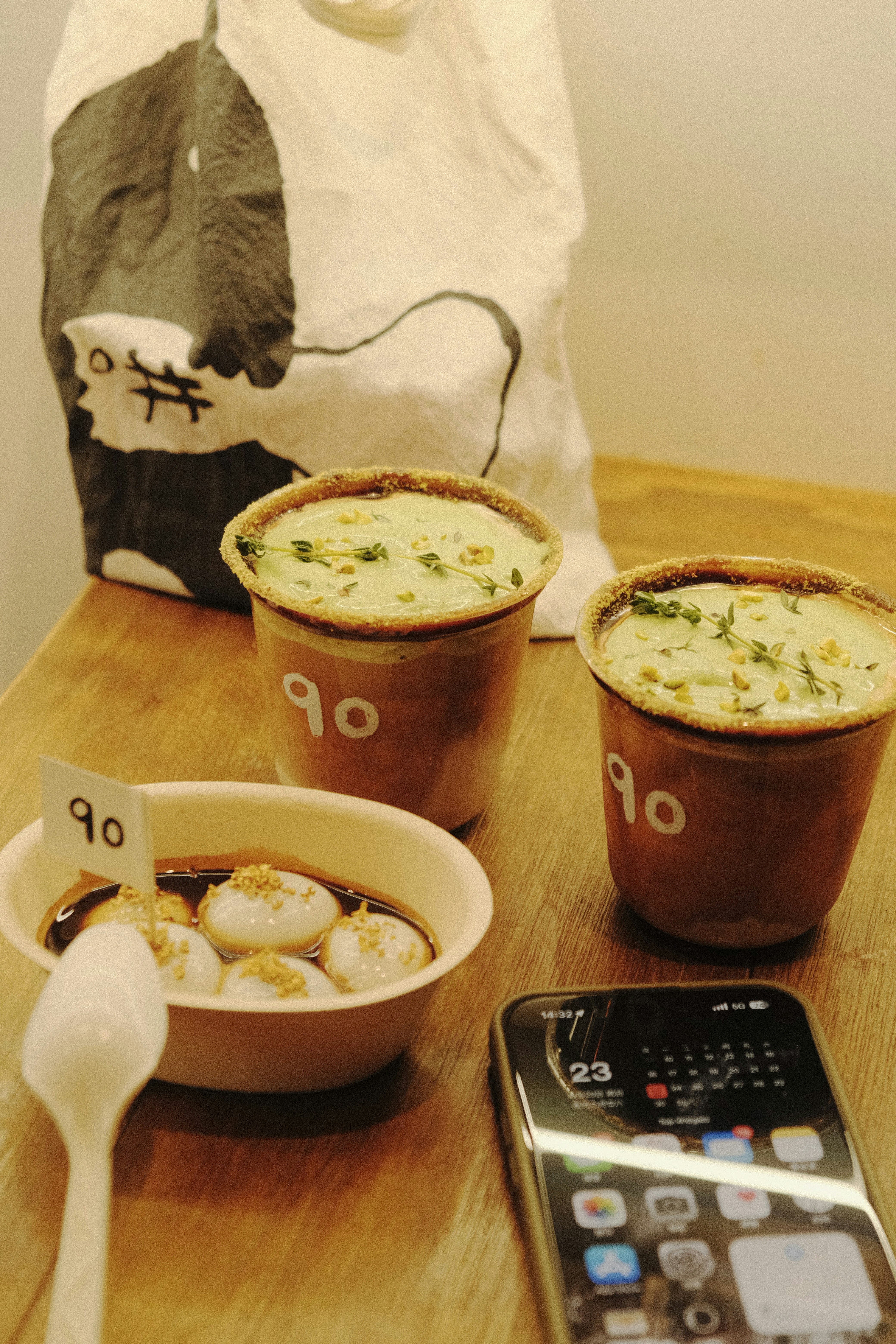 Two clay pots filled with creamy dessert sit beside a bowl of sweet dumplings and a smartphone on a wooden table. A stylish bag adds a touch of modernity.