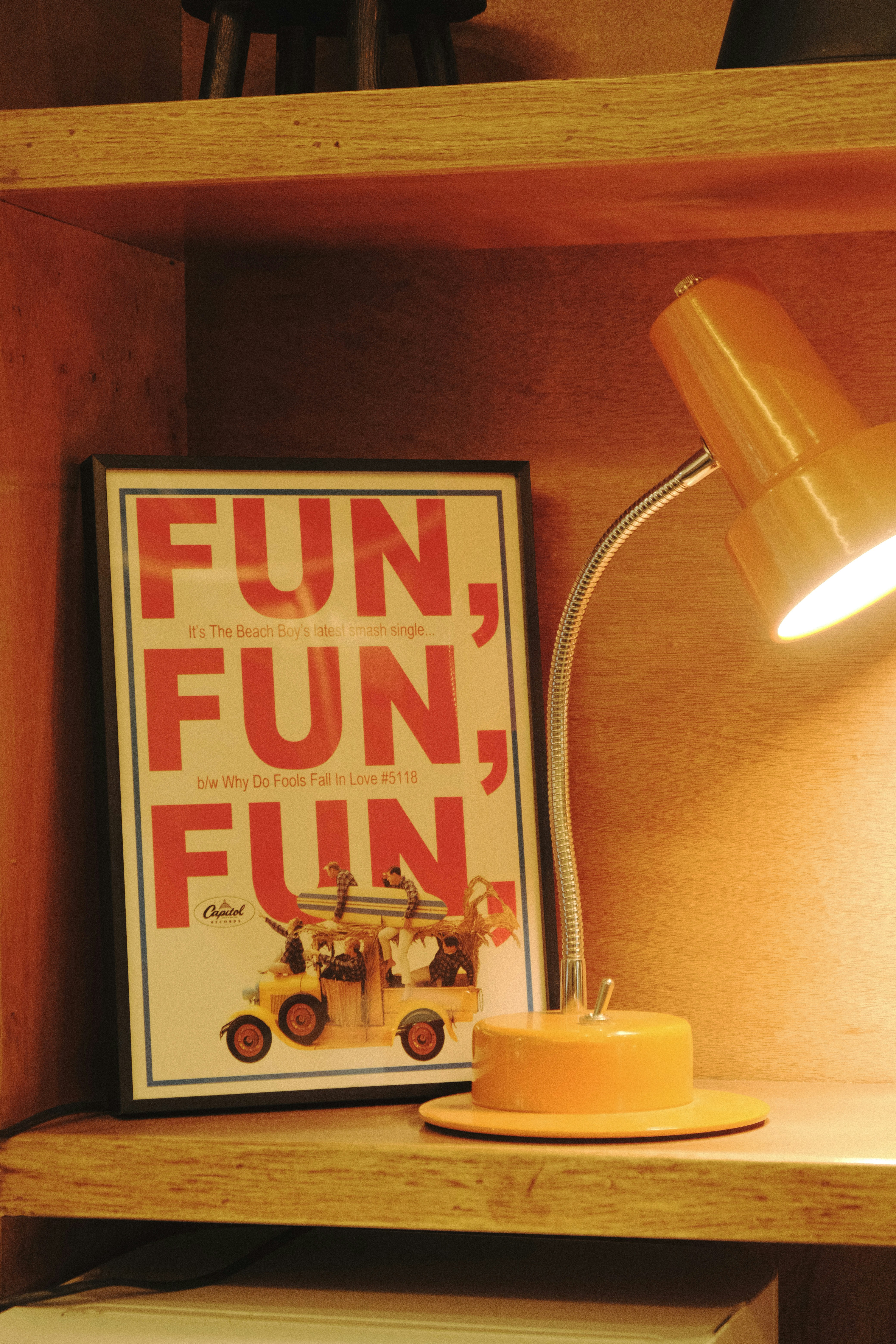 Framed vintage poster on a wood shelf beside a yellow desk lamp. The bold red typography and retro imagery give the scene a warm, nostalgic vibe.