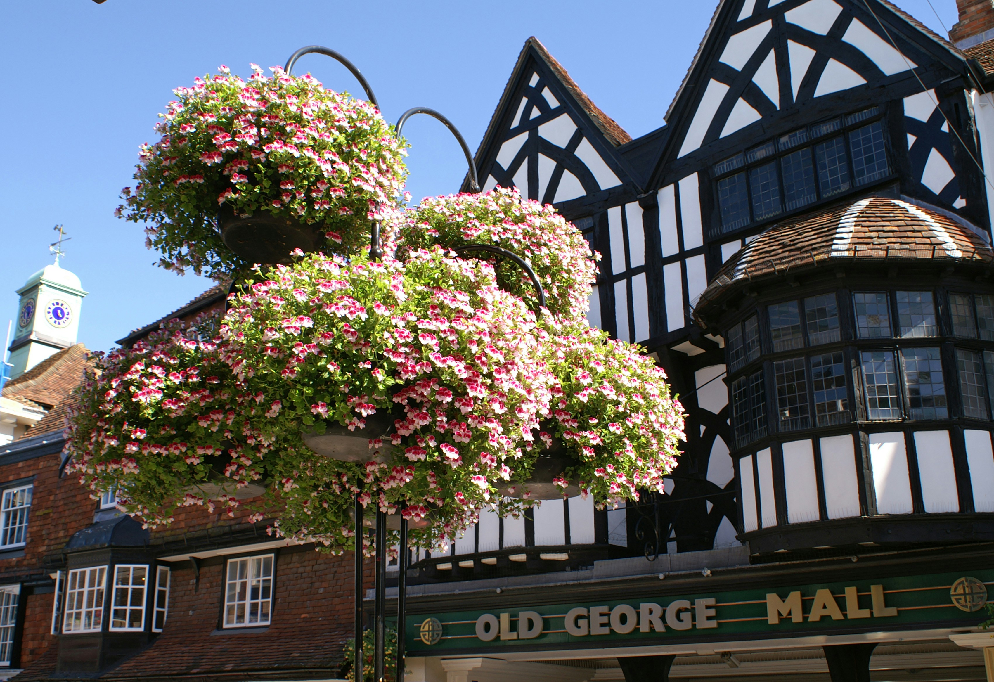 A street scene showcasing a Tudor-style timber-framed facade with vibrant pink hanging baskets above the Old George Mall sign.