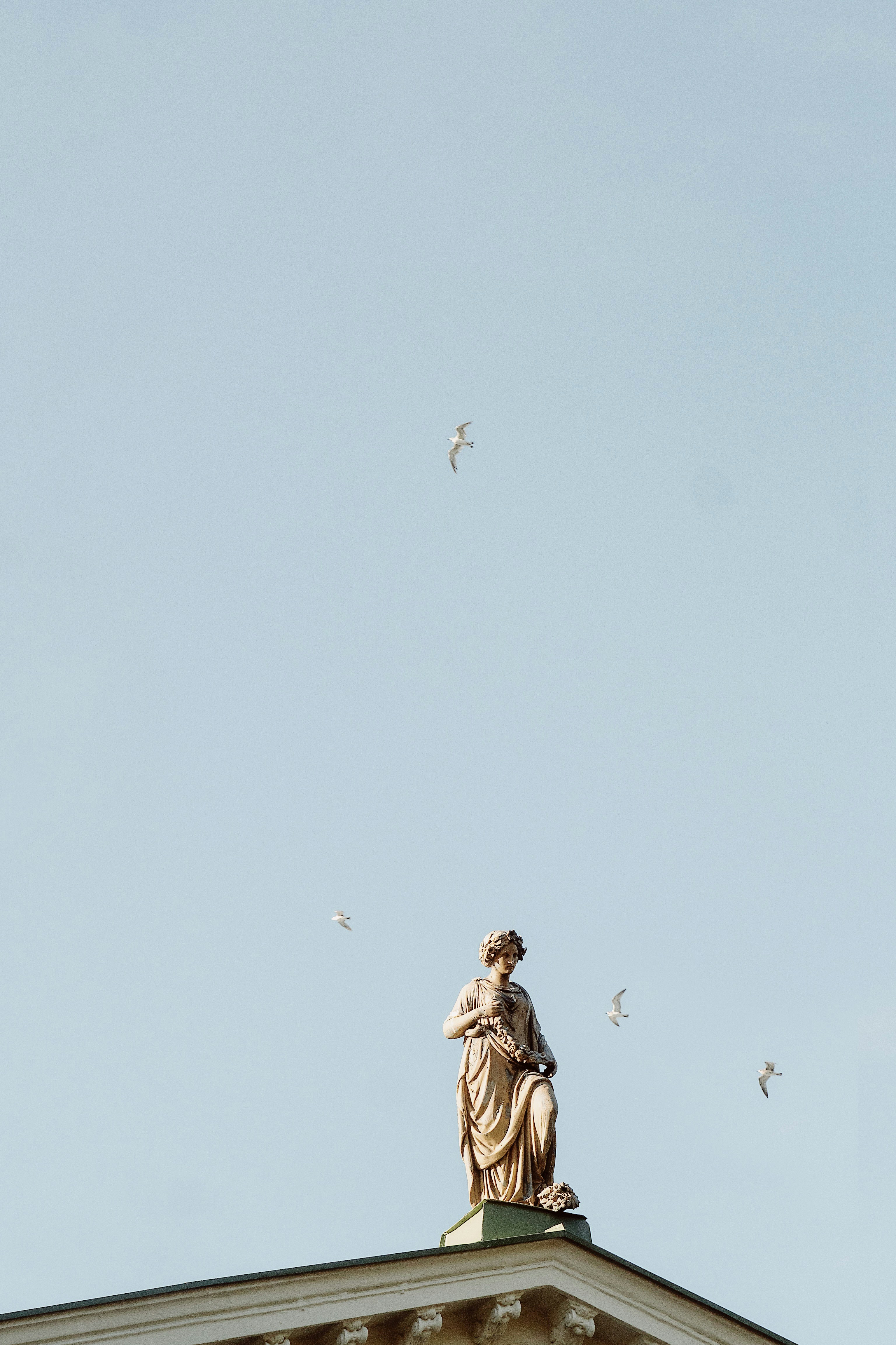 Classical statue perched atop a rooftop against a pale blue sky. Several doves drift nearby, adding a sense of motion to the serene scene.