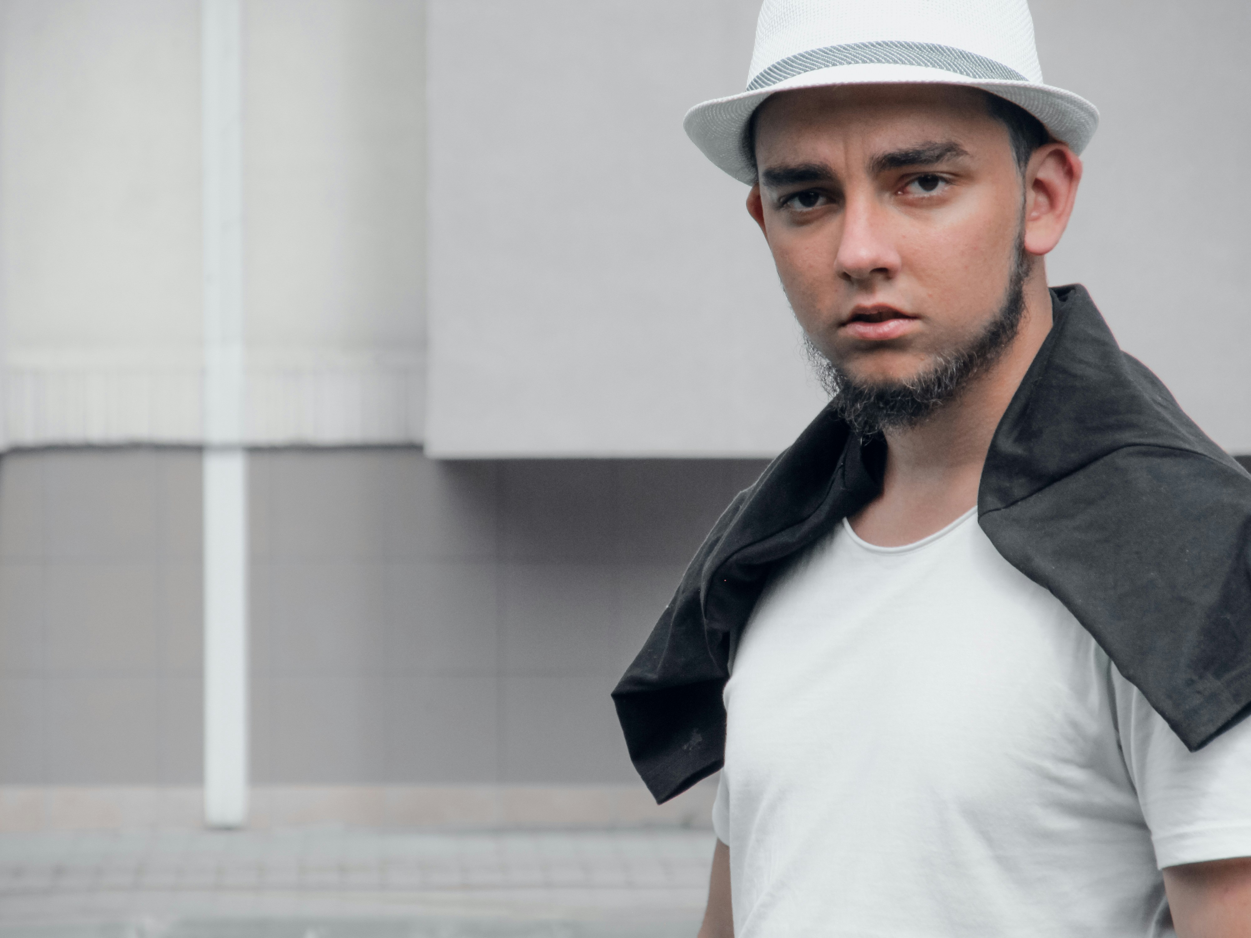 Young man in a white hat and casual attire poses confidently against a minimalist backdrop.