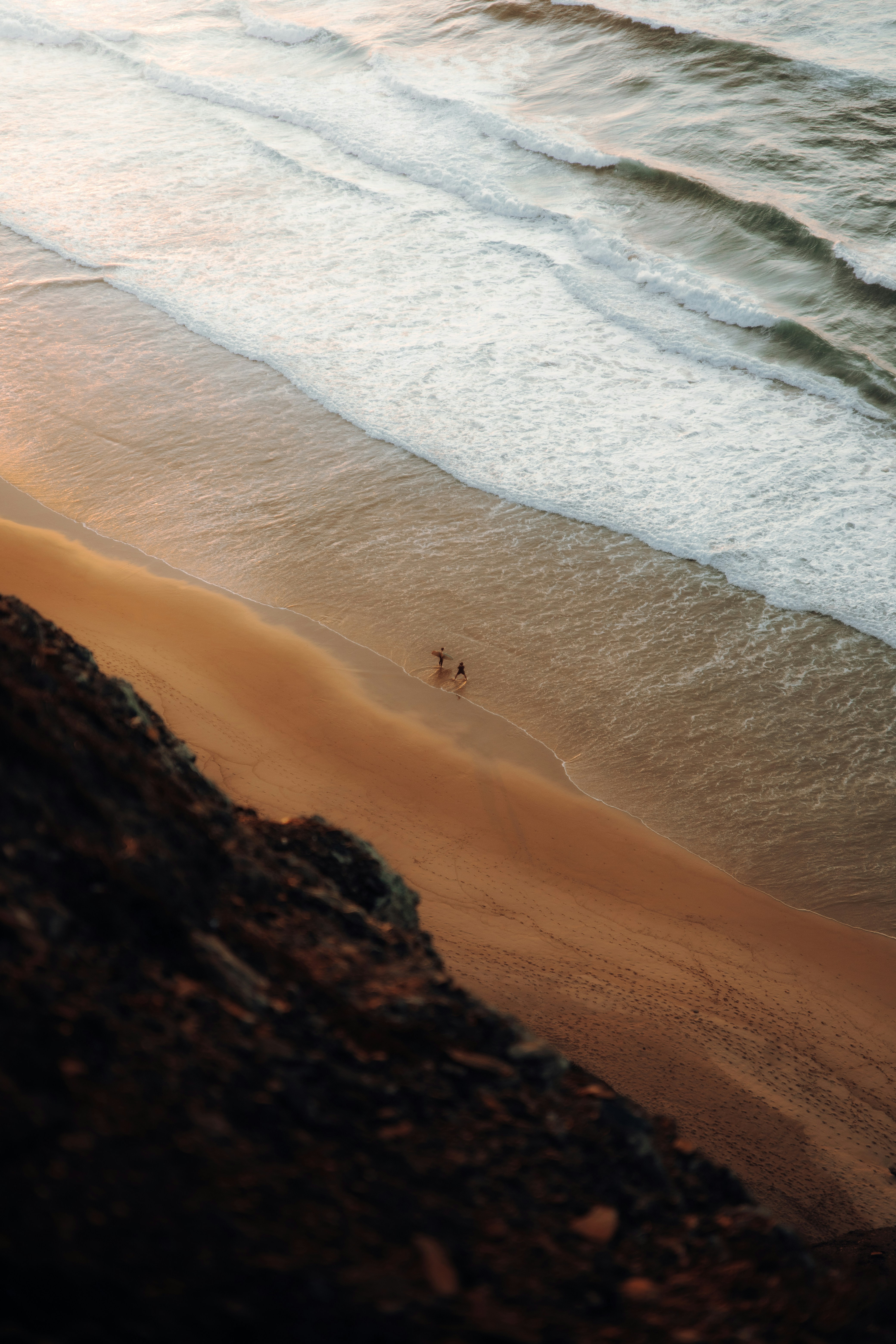 Two figures walking hand in hand on a sandy beach as waves gently lap at the shore during sunset.