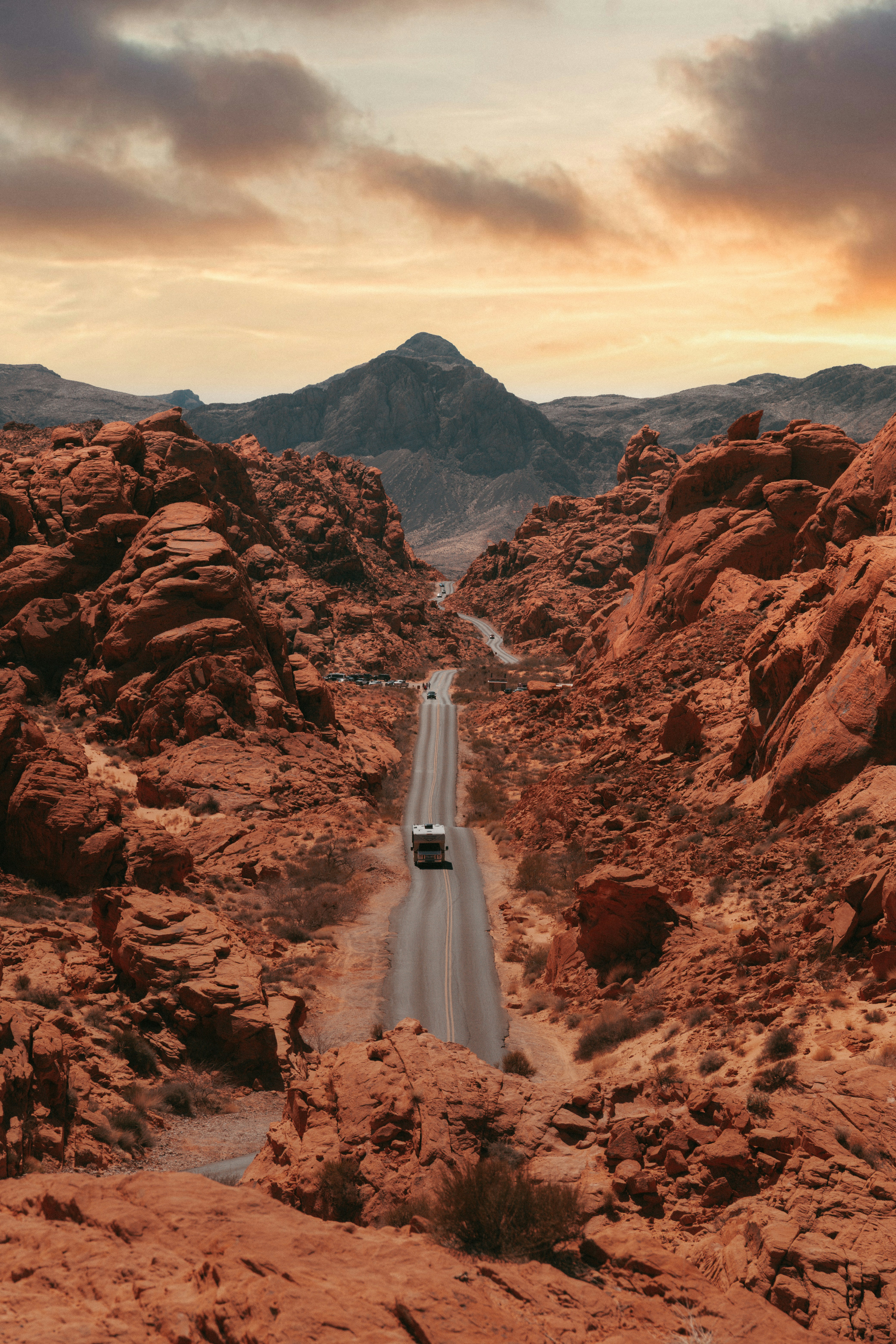 A lone van travels a narrow road carved through towering red rock formations in a desert canyon at dusk.