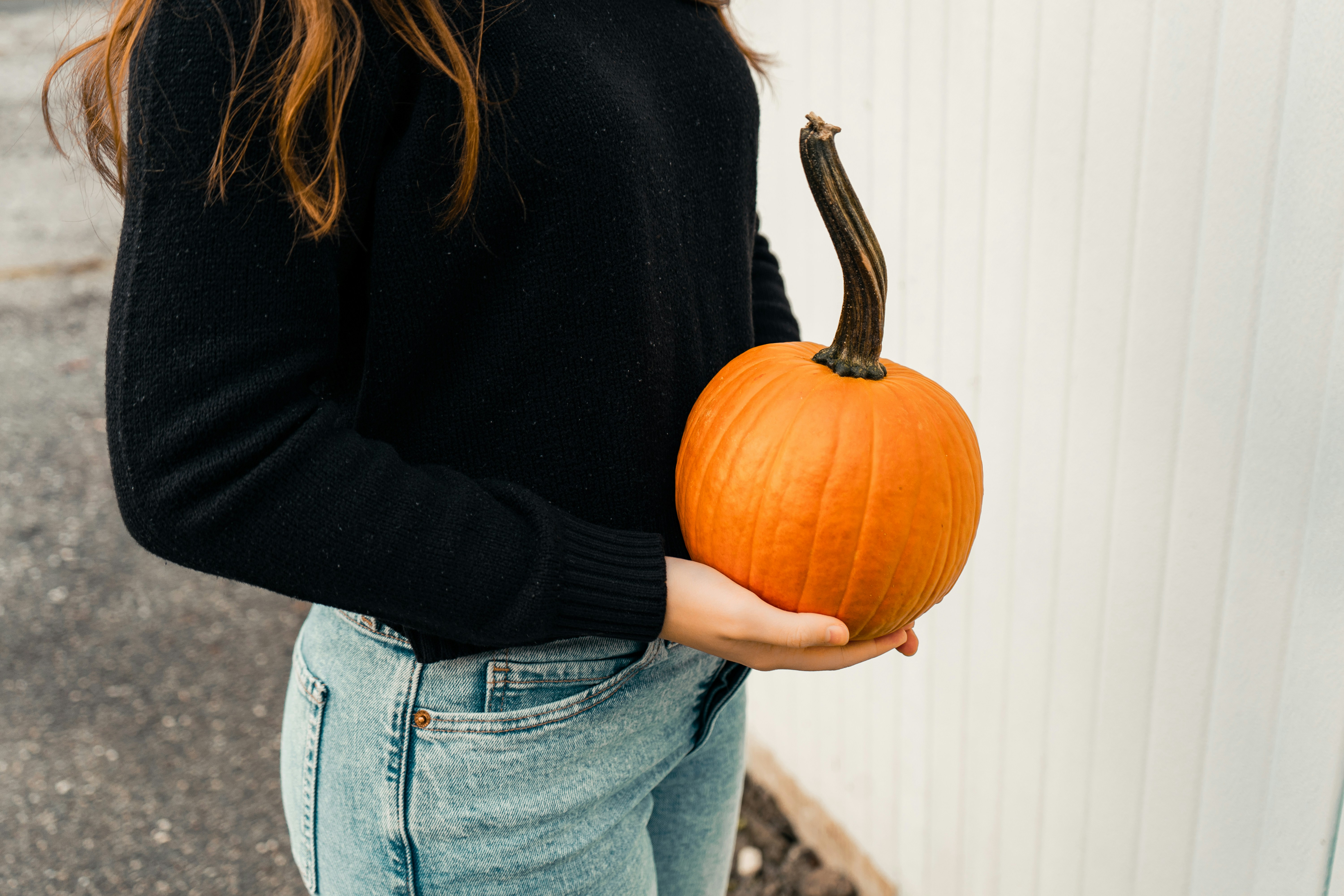 Person in a black sweater holding a small pumpkin against a pale background.