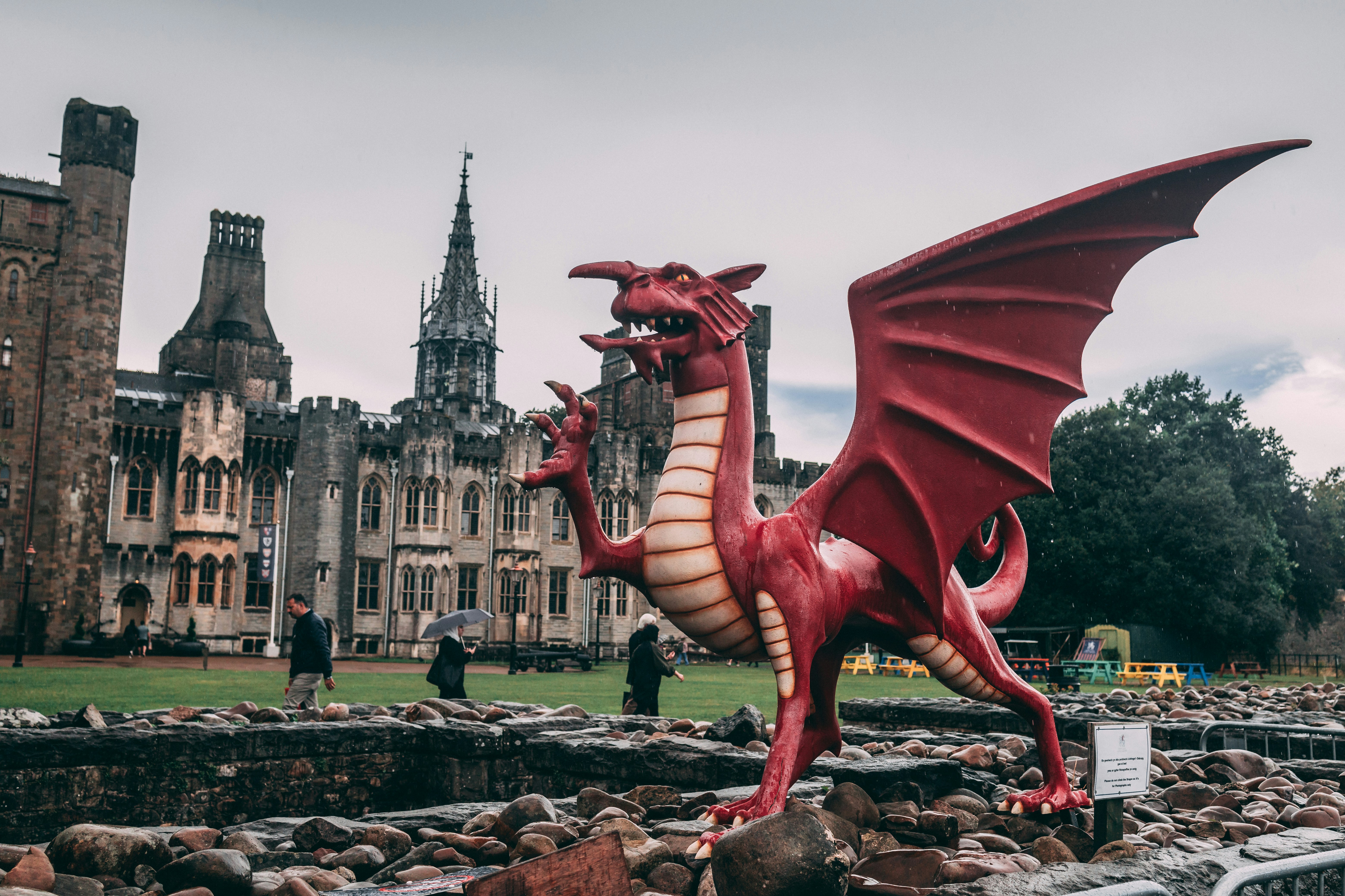 Vibrant red dragon sculpture stands proudly in front of historic architecture, symbolizing Welsh culture. Visitors explore the grounds nearby.