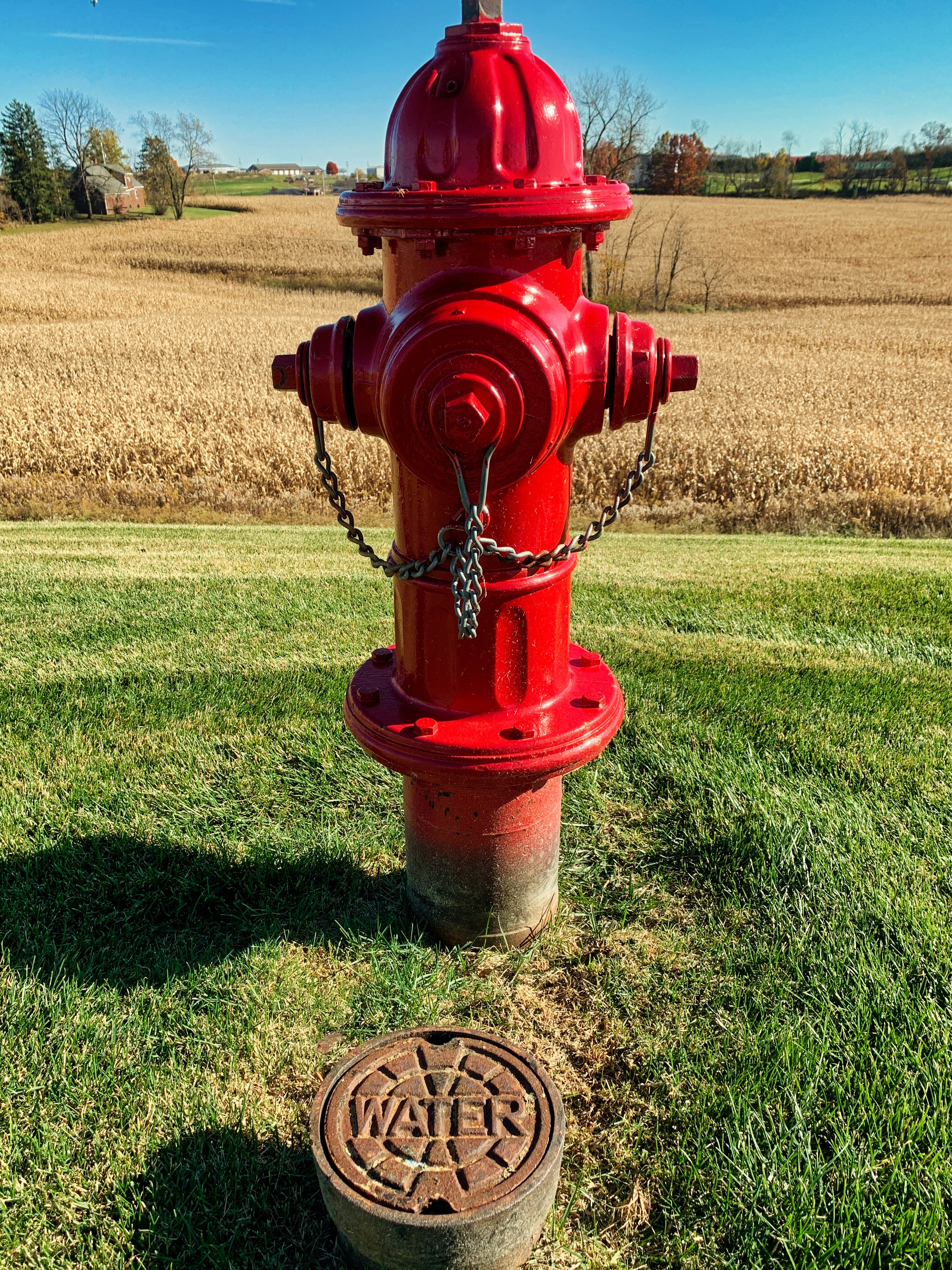 Bright red fire hydrant standing prominently on a grassy knoll, overlooking golden fields. Water access cover visible in the foreground.