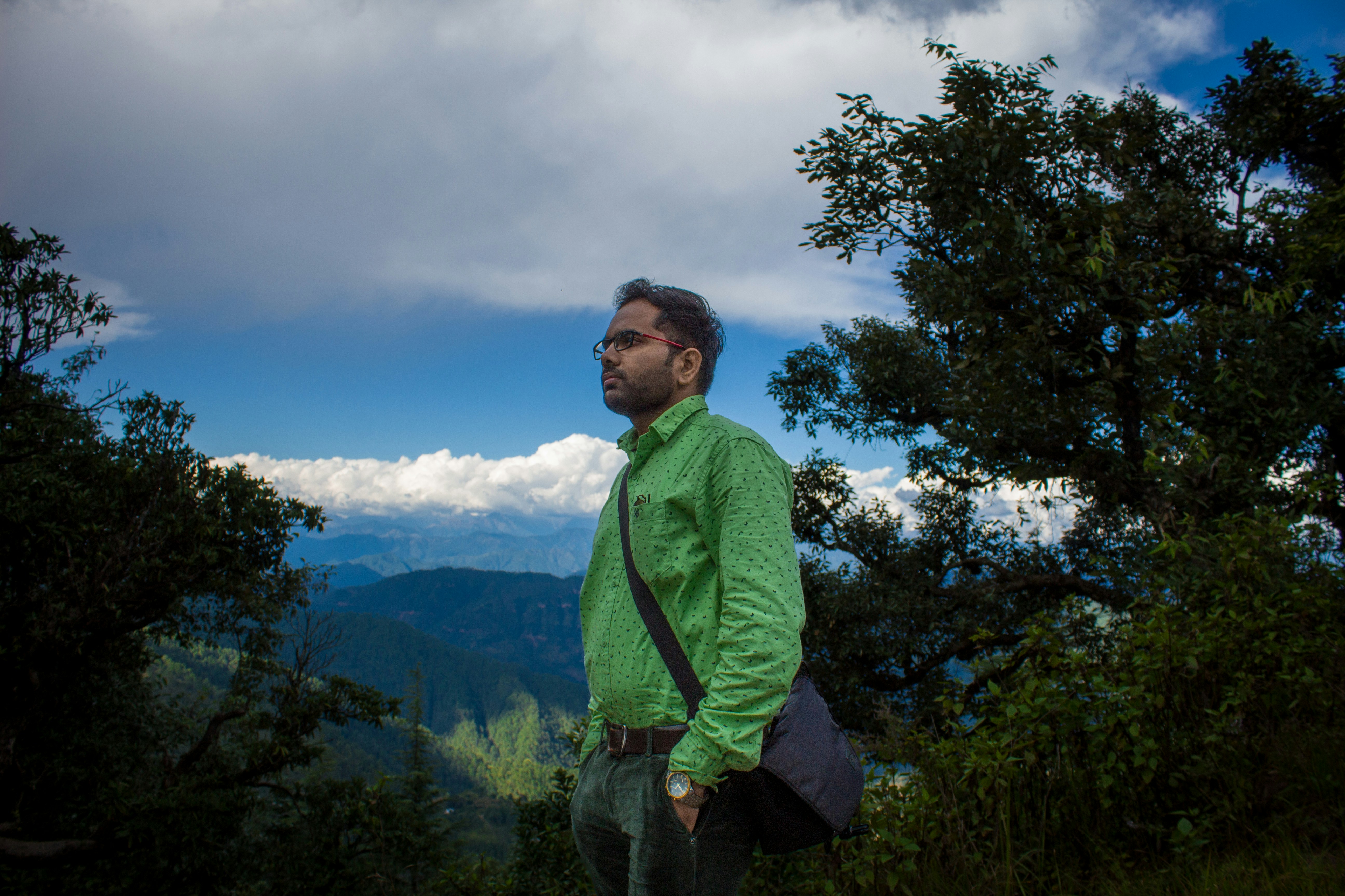 Man in a green shirt standing on a mountain viewpoint surrounded by lush trees and distant peaks under a cloudy sky.
