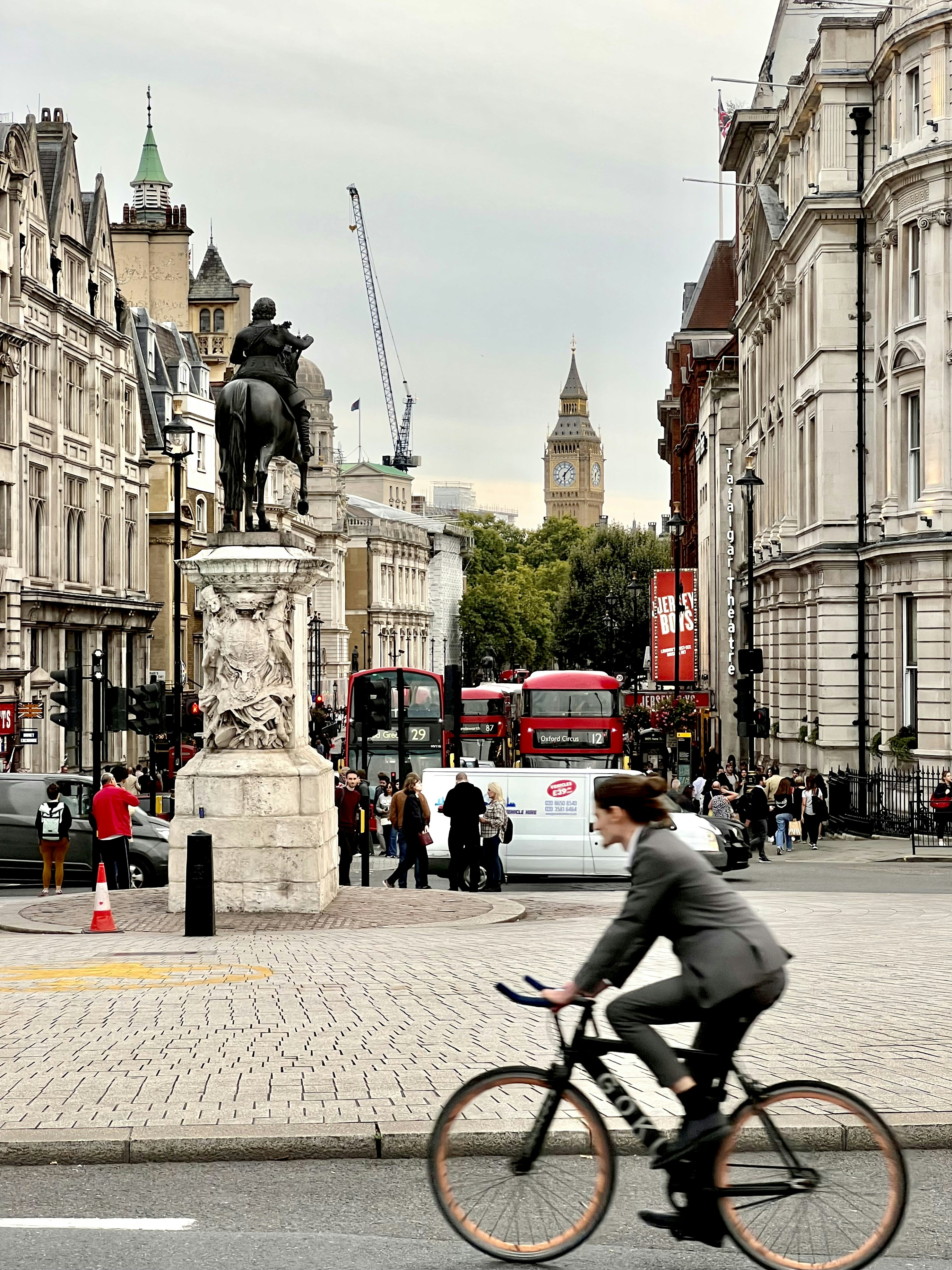 Cyclist passing through a bustling London street with iconic red buses and Big Ben in the background.
