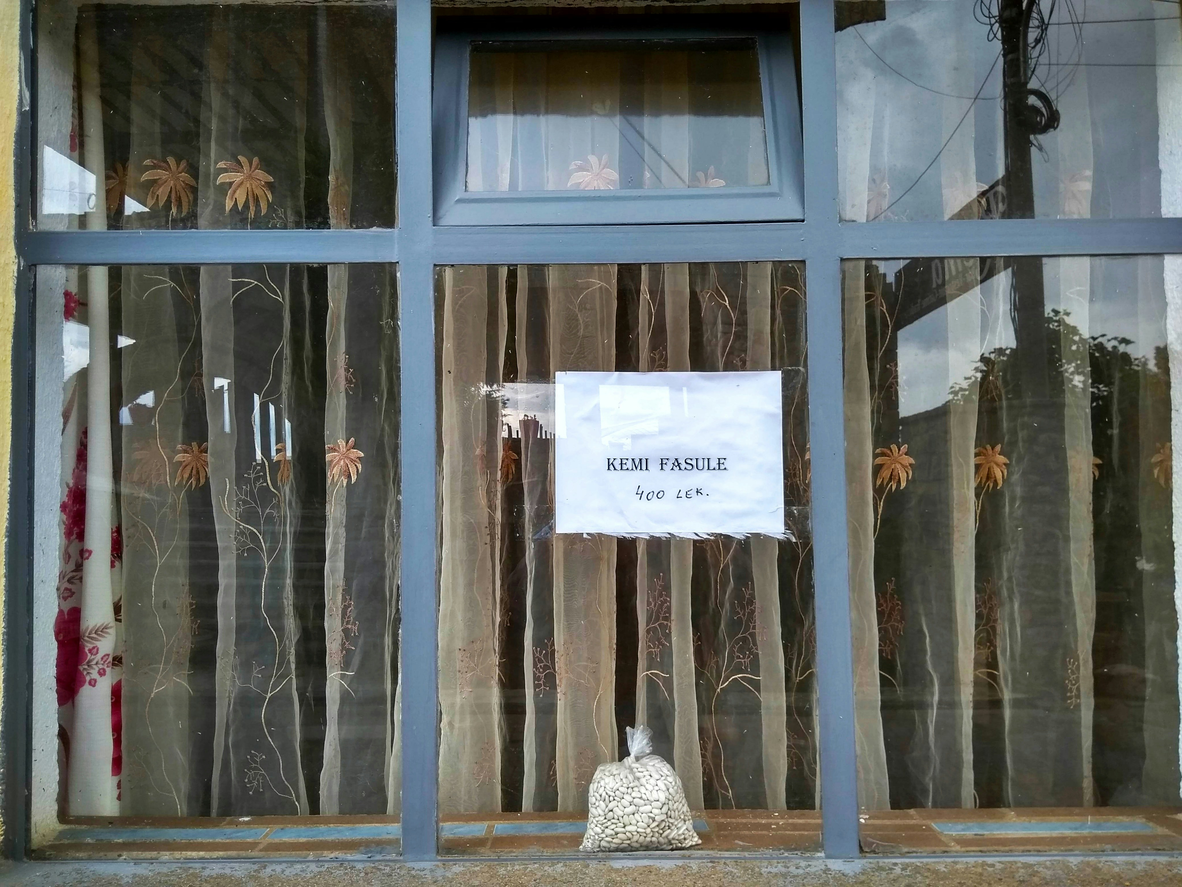 Storefront with large windows reflecting the street, adorned with simple curtains and a central sign.
