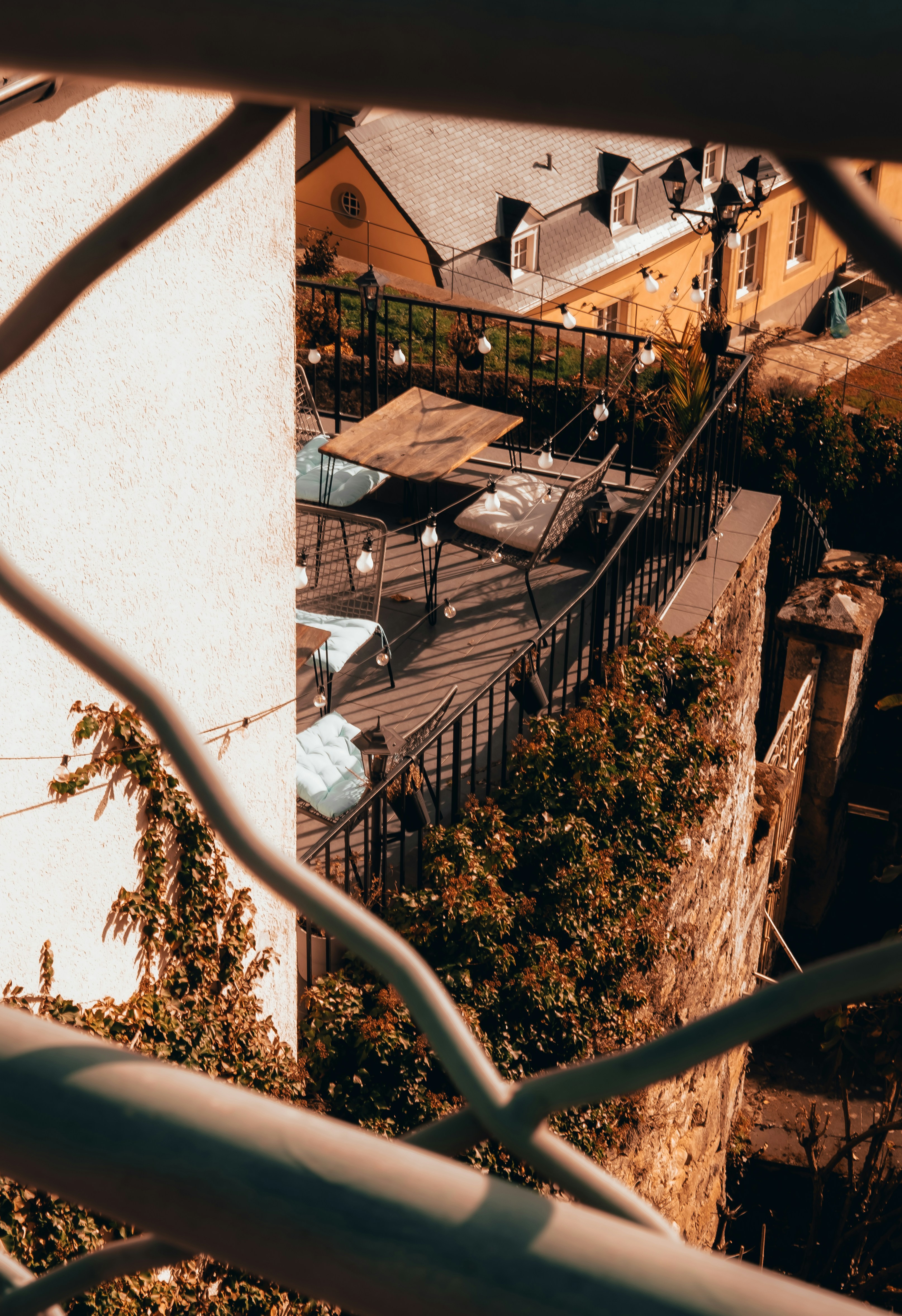 Balcony terrace adorned with lounge chairs and string lights, surrounded by greenery and rustic architecture.