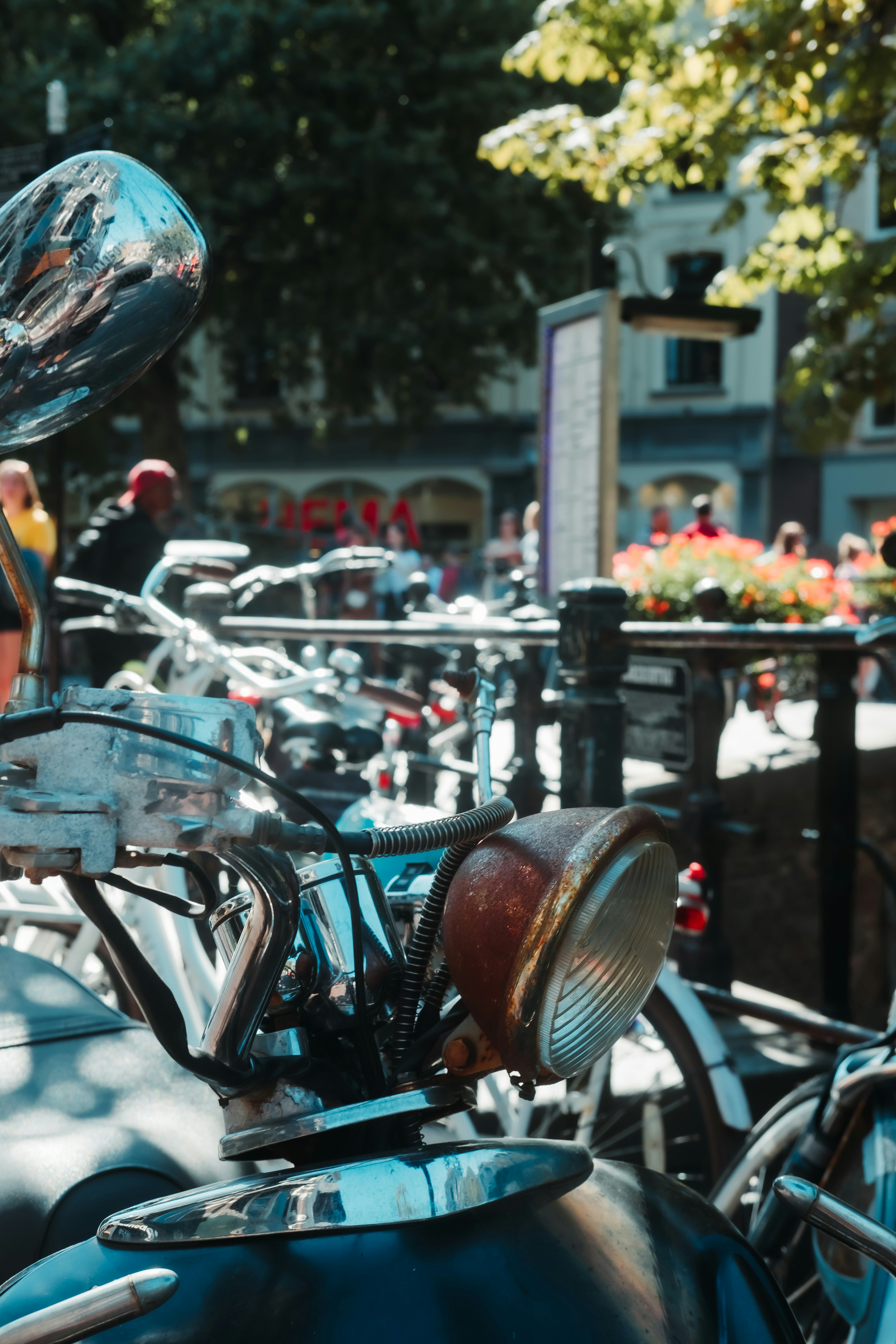 Close-up of a vintage motorcycle headlamp with gleaming chrome details, surrounded by bicycles and vibrant flowers in the background.
