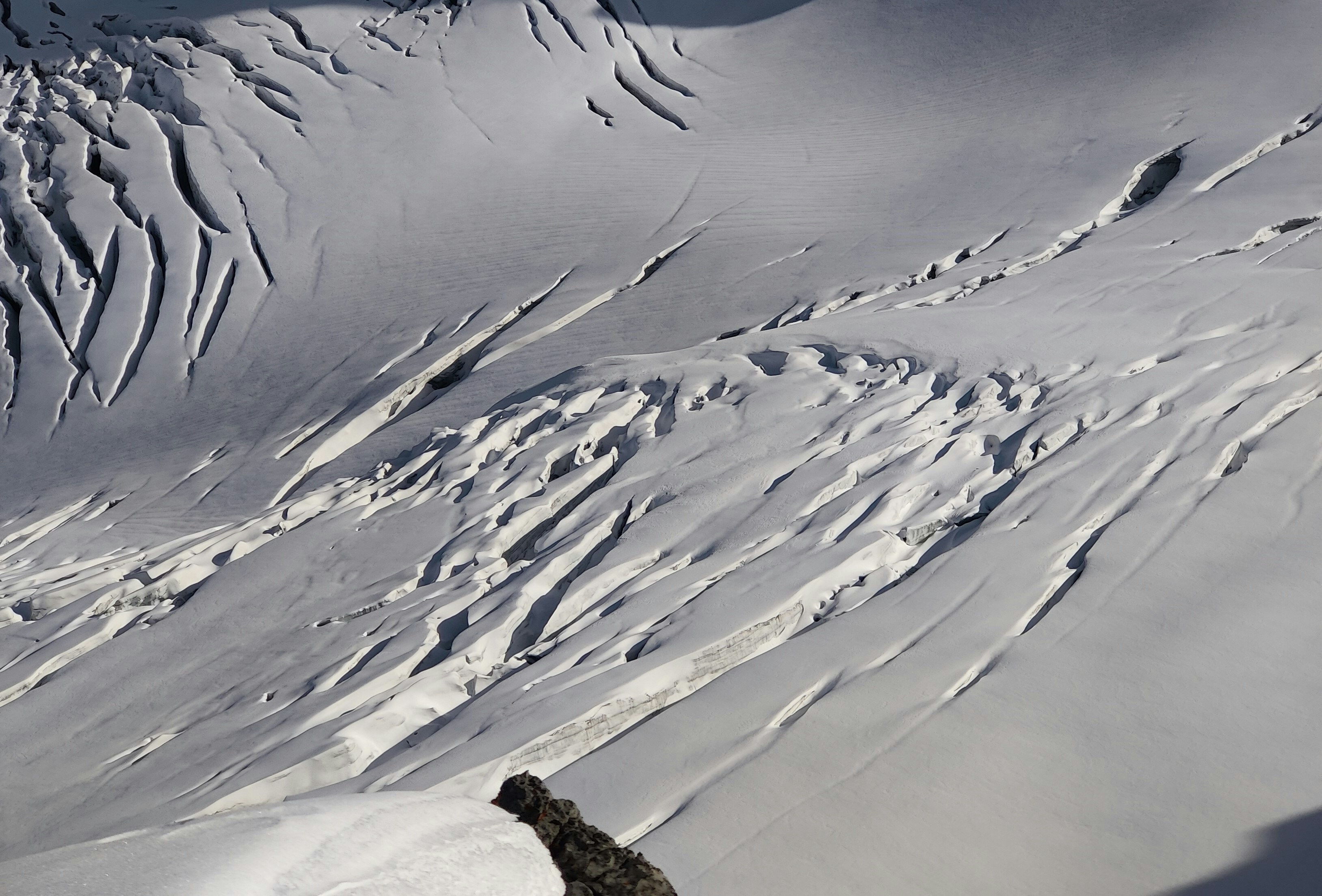 Snow-covered slope with wind-carved ridges and a solitary rock at the lower edge, forming a stark diagonal texture.