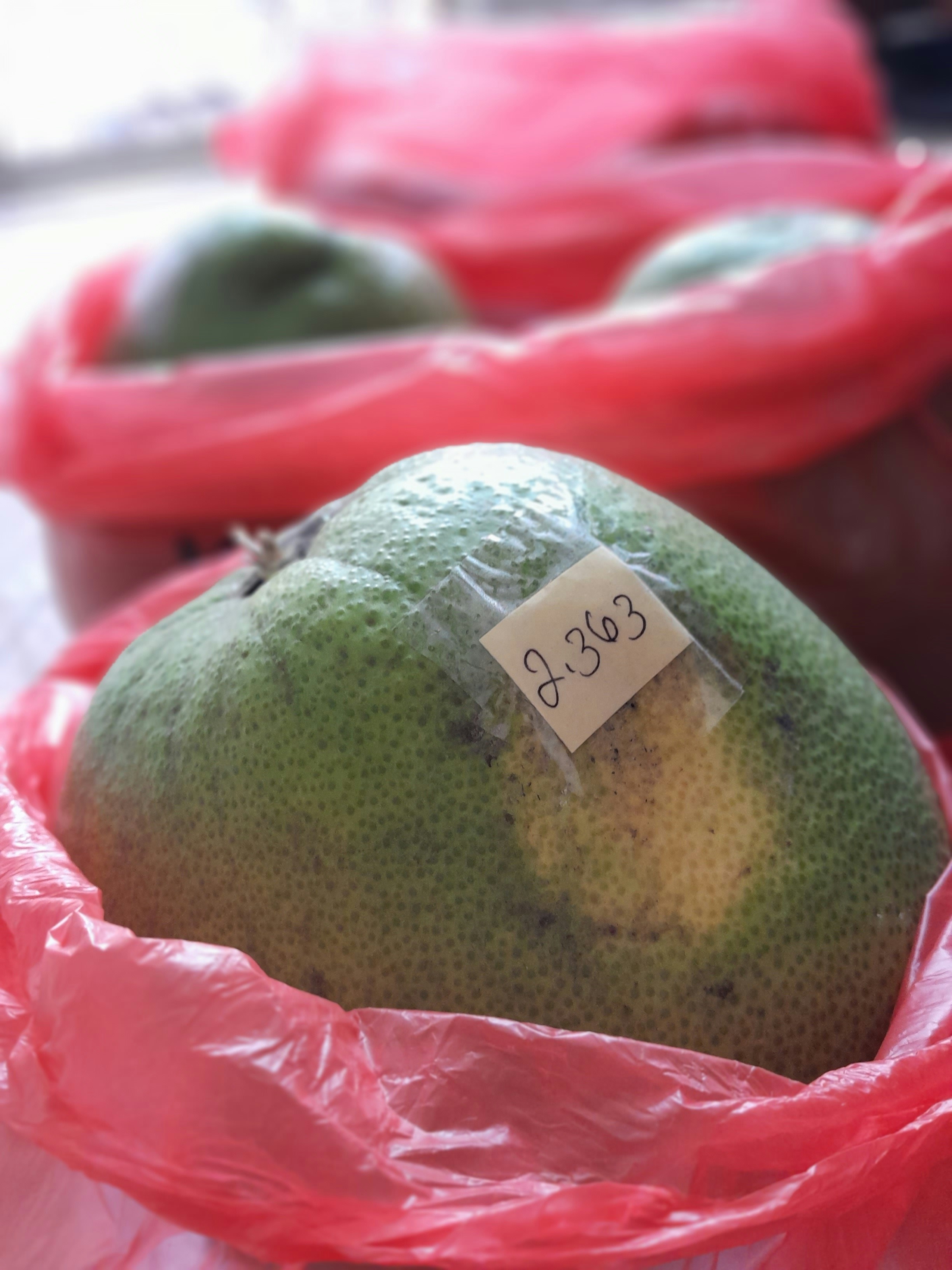 Close-up photograph of a green pomelo in a pink plastic bag, with a small sticker reading '8-863'.