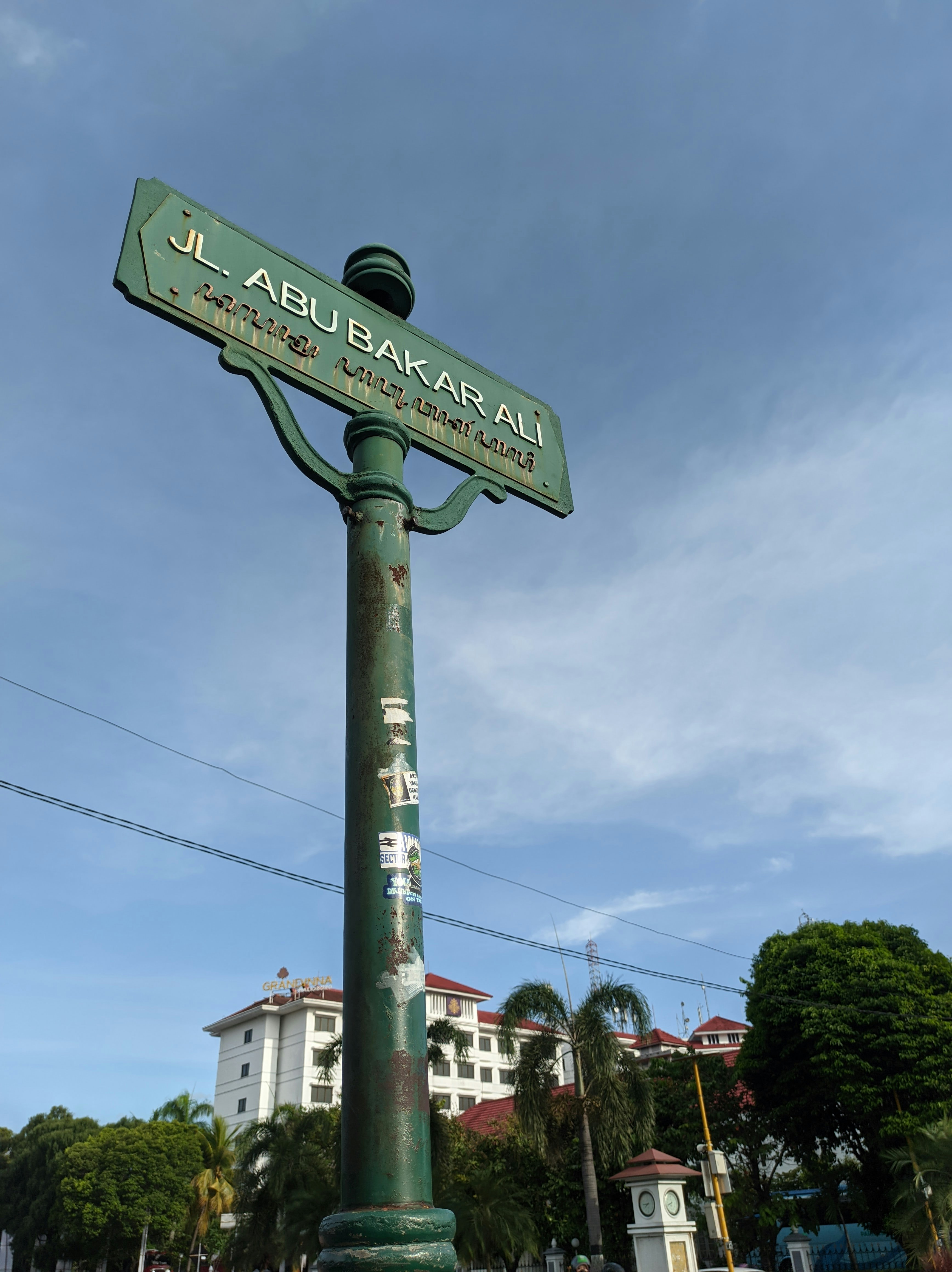 Green street sign indicating 'Jl. Abu Bakar Ali' with a backdrop of palm trees and a clear blue sky.