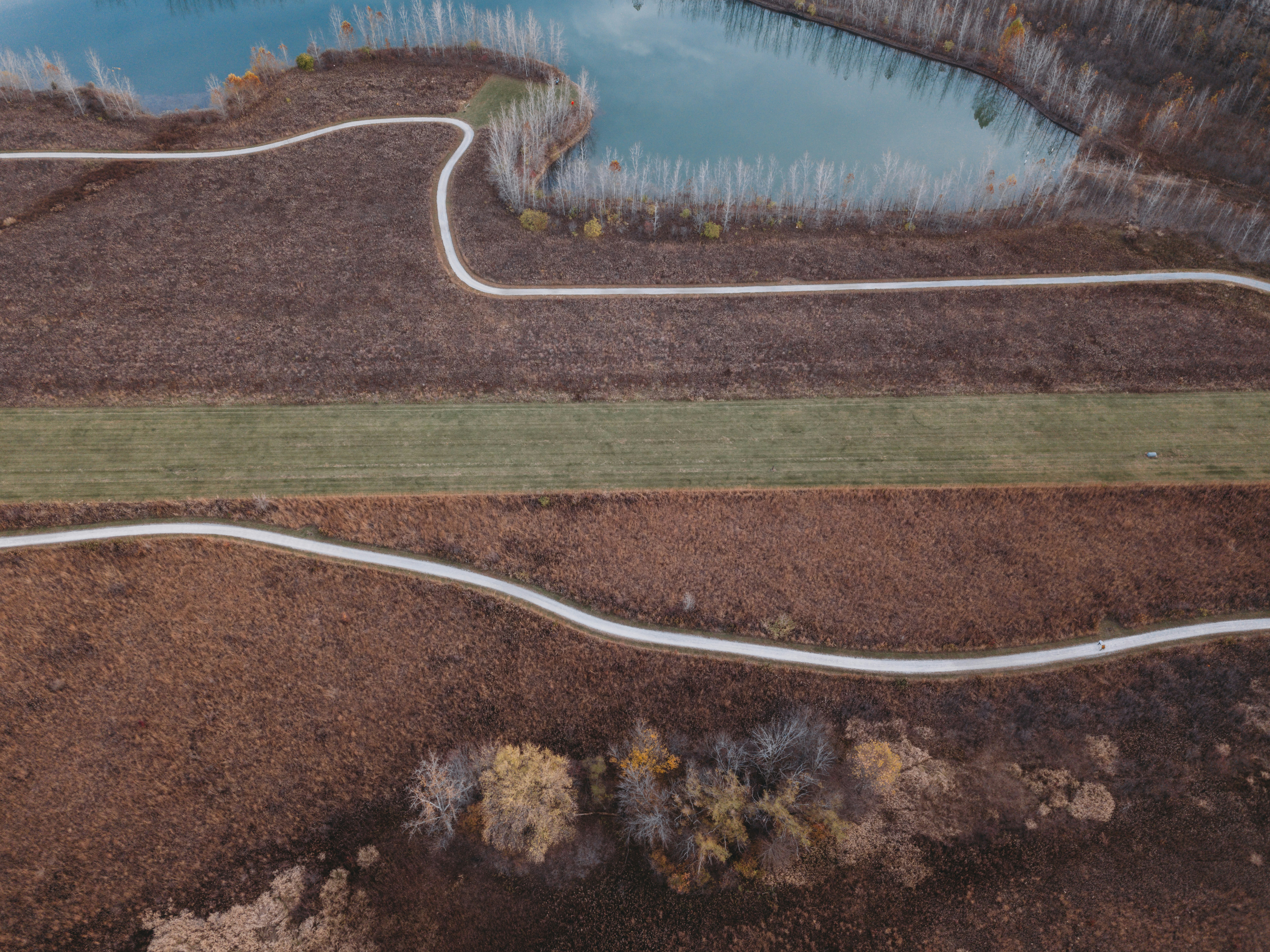 Aerial view of winding paths and a serene lake surrounded by autumn foliage, showcasing the seasonal transition in a tranquil landscape.