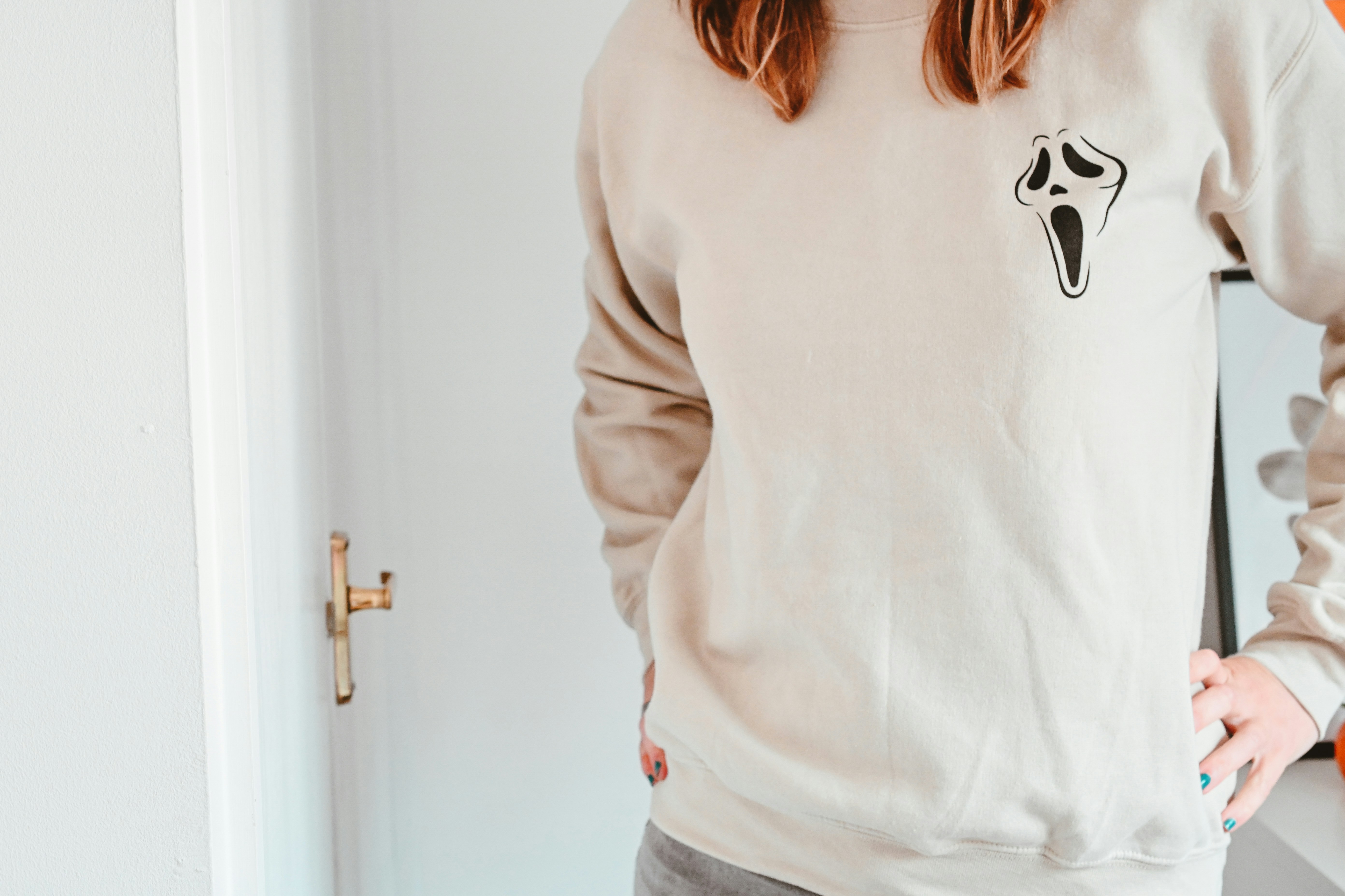 A person wearing a beige sweatshirt featuring the Ghostface logo stands in a softly lit room, exuding a blend of casual style and pop culture reference.