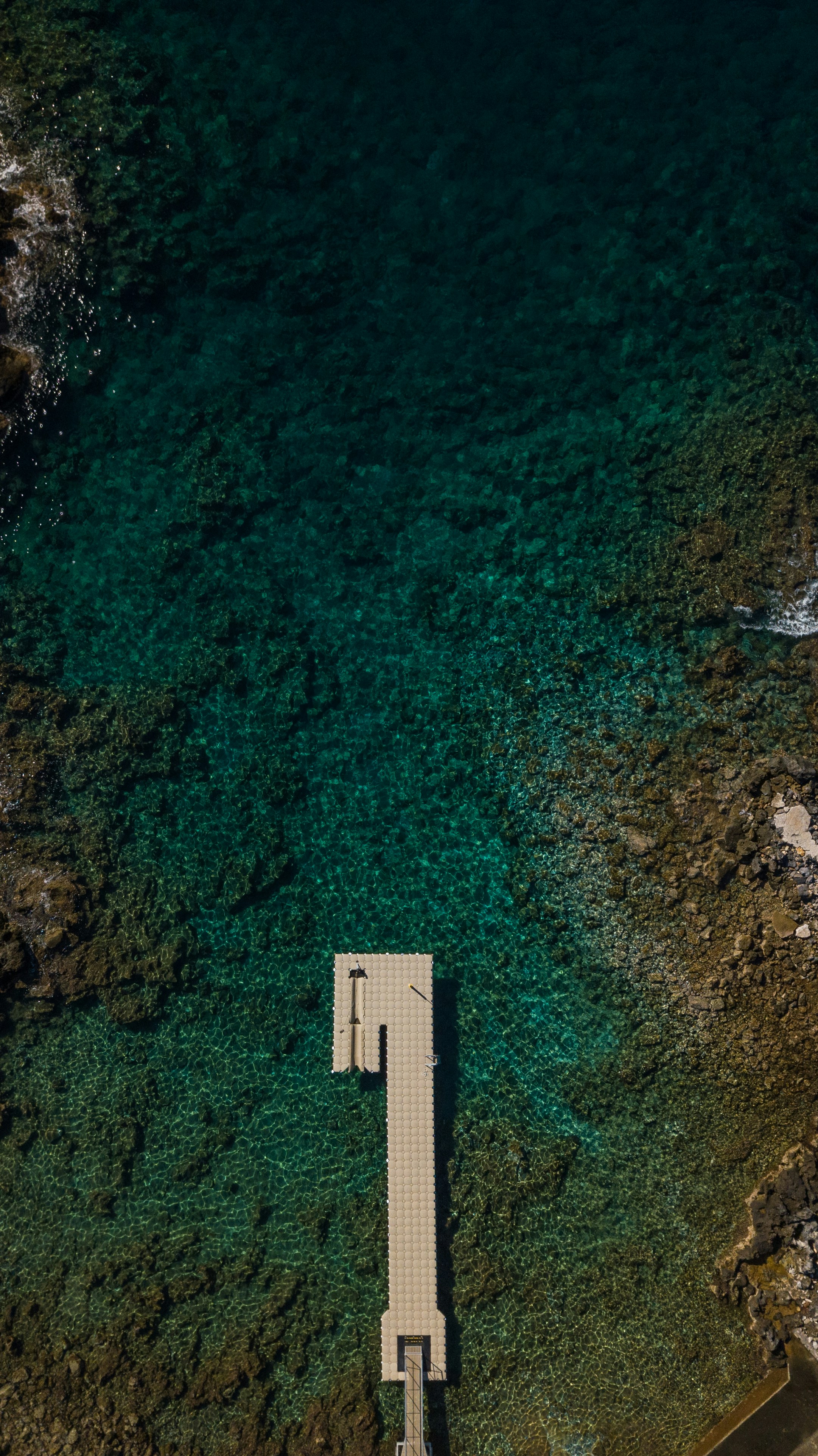A wooden dock stretching into crystal-clear waters, surrounded by rocky formations. The aerial perspective highlights the vibrant turquoise hues of the sea.