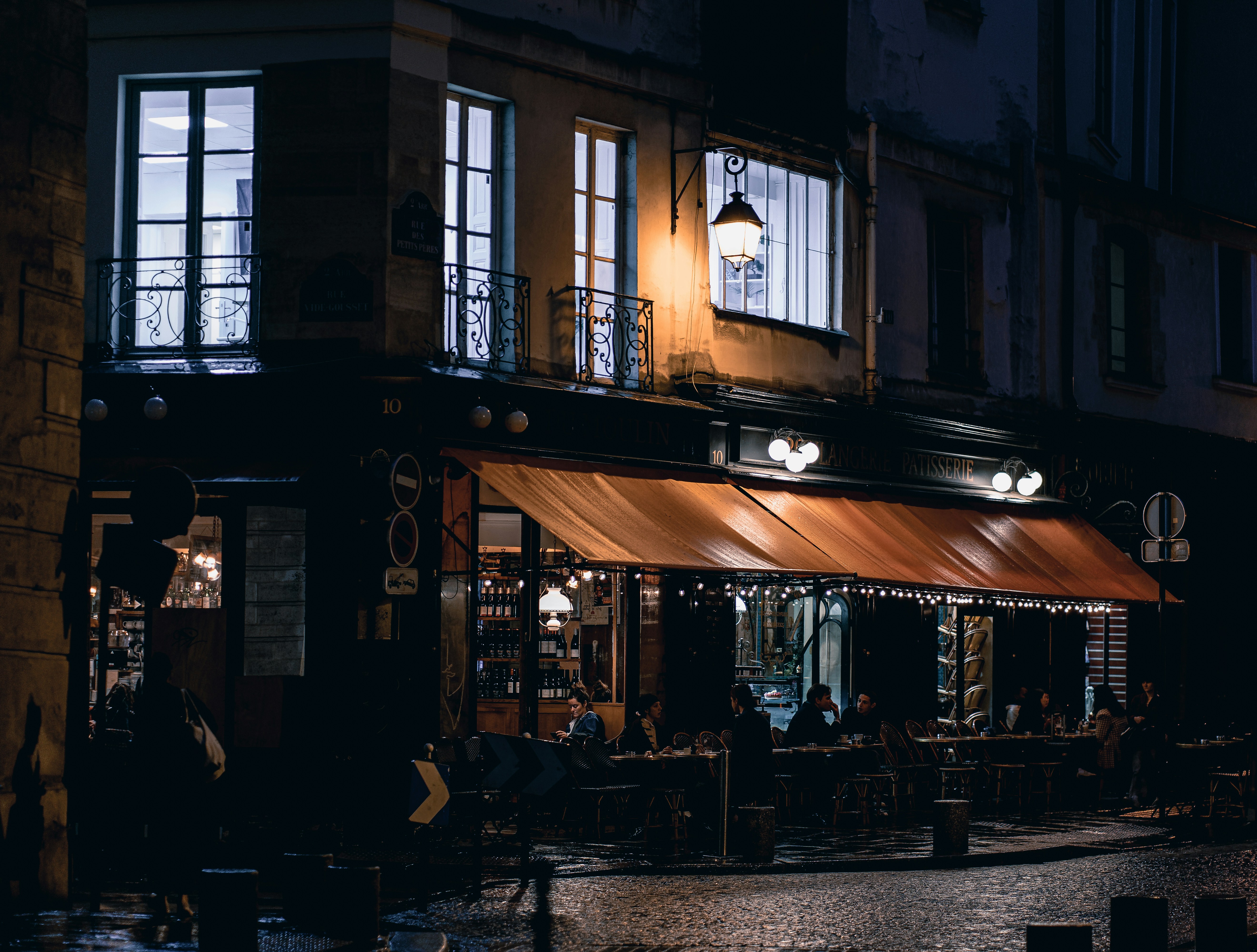 Cozy bistro illuminated by warm lights, bustling with patrons under a soft orange awning. The scene captures the essence of urban nightlife.