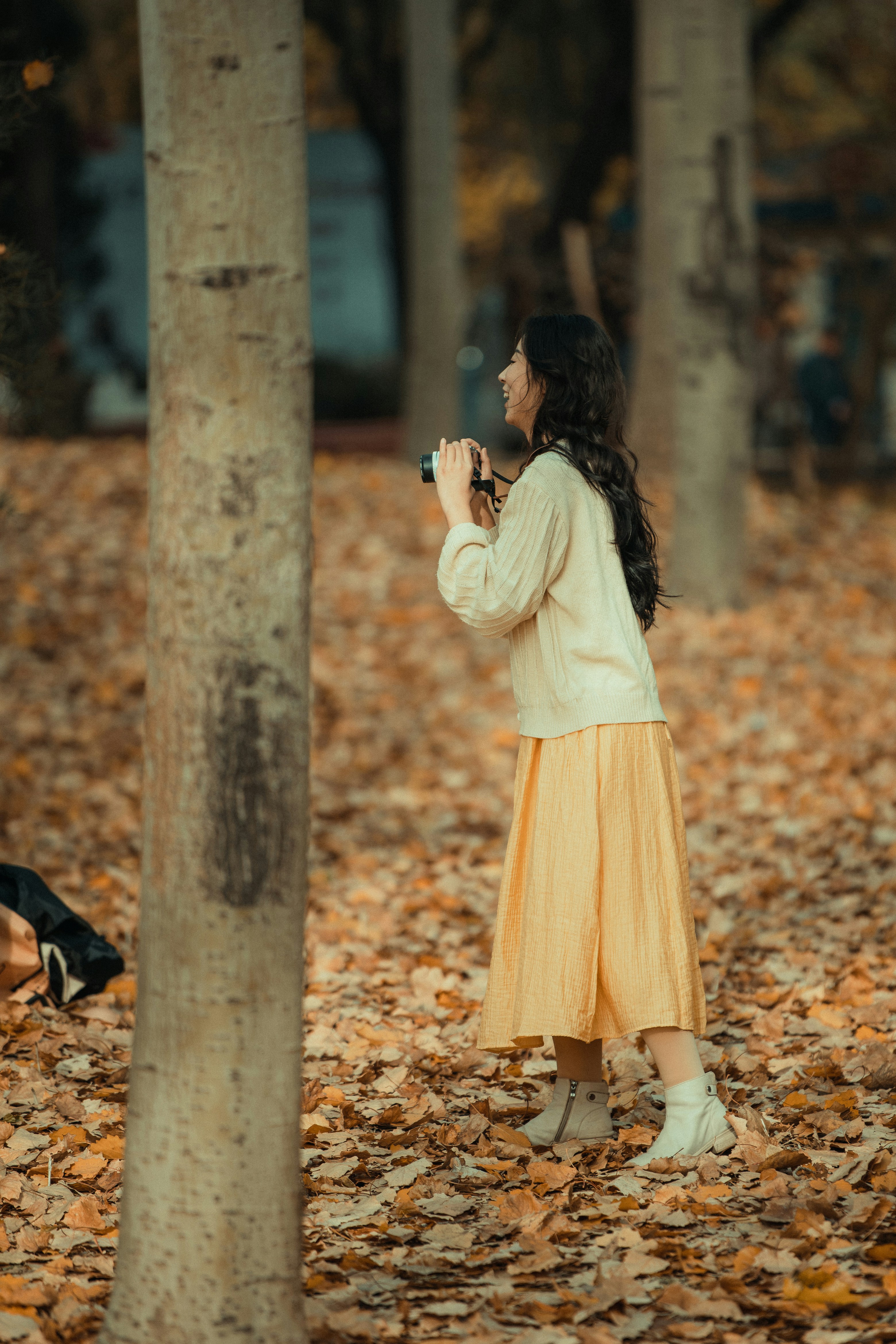 A woman with a camera walks through a park strewn with orange leaves; the focus is on her as she aims the camera, capturing a candid autumn moment. Photograph type: lifestyle portrait.