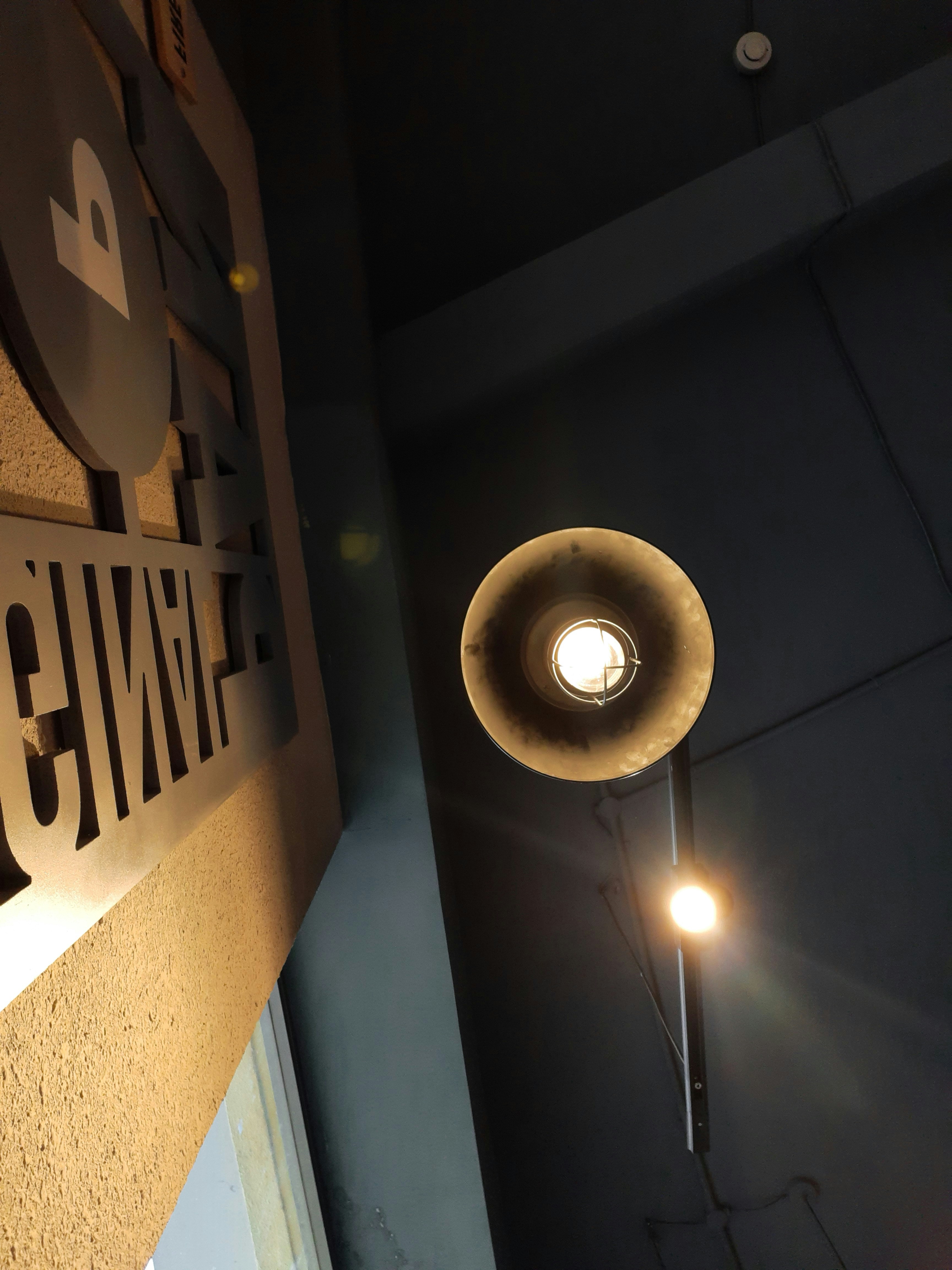A single circular ceiling lamp casts a warm halo on a dark ceiling, with a slim vertical light fixture and partial signage visible on the left.