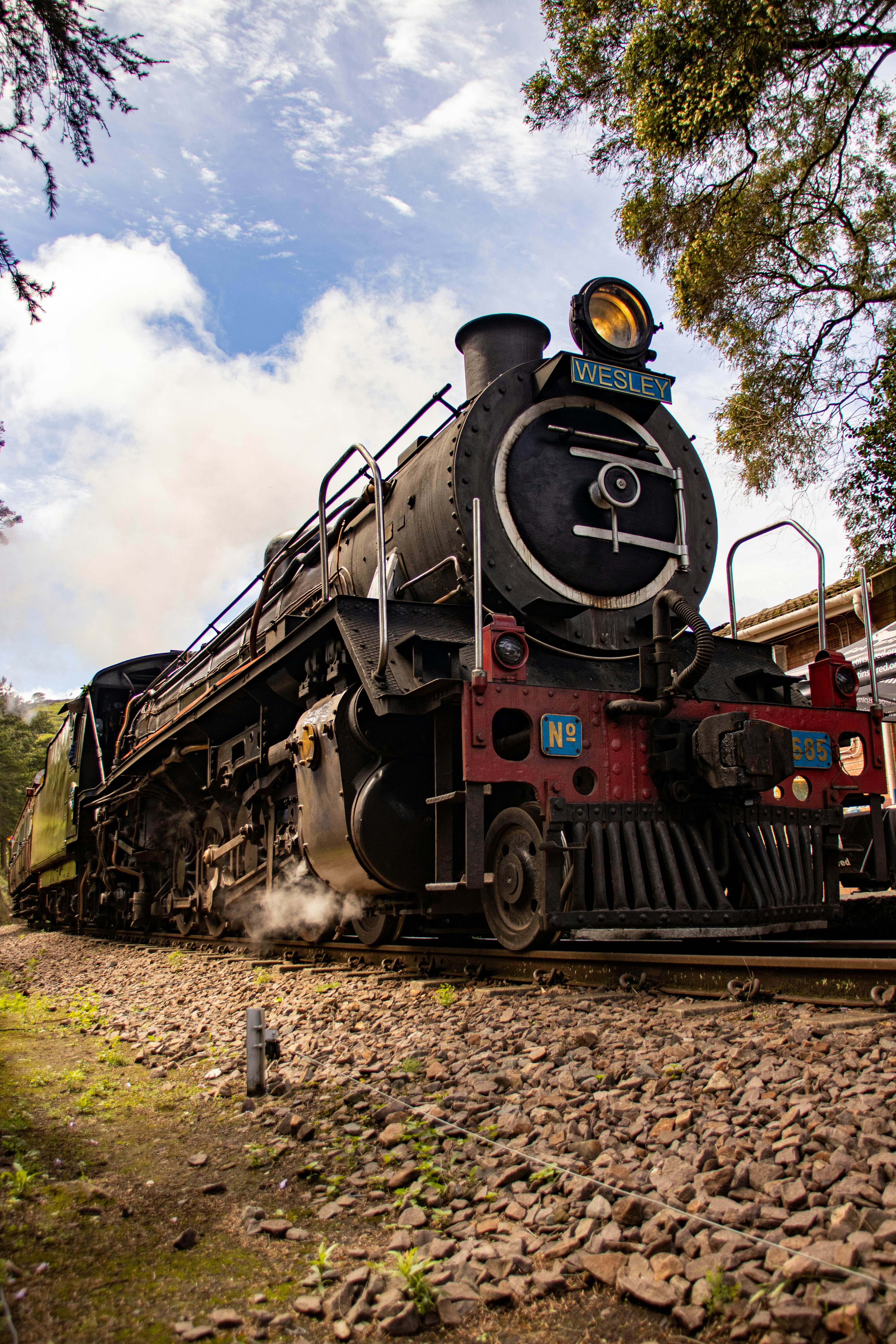 Vintage steam locomotive named Wesley, showcasing intricate details and billowing steam against a backdrop of trees and blue sky.
