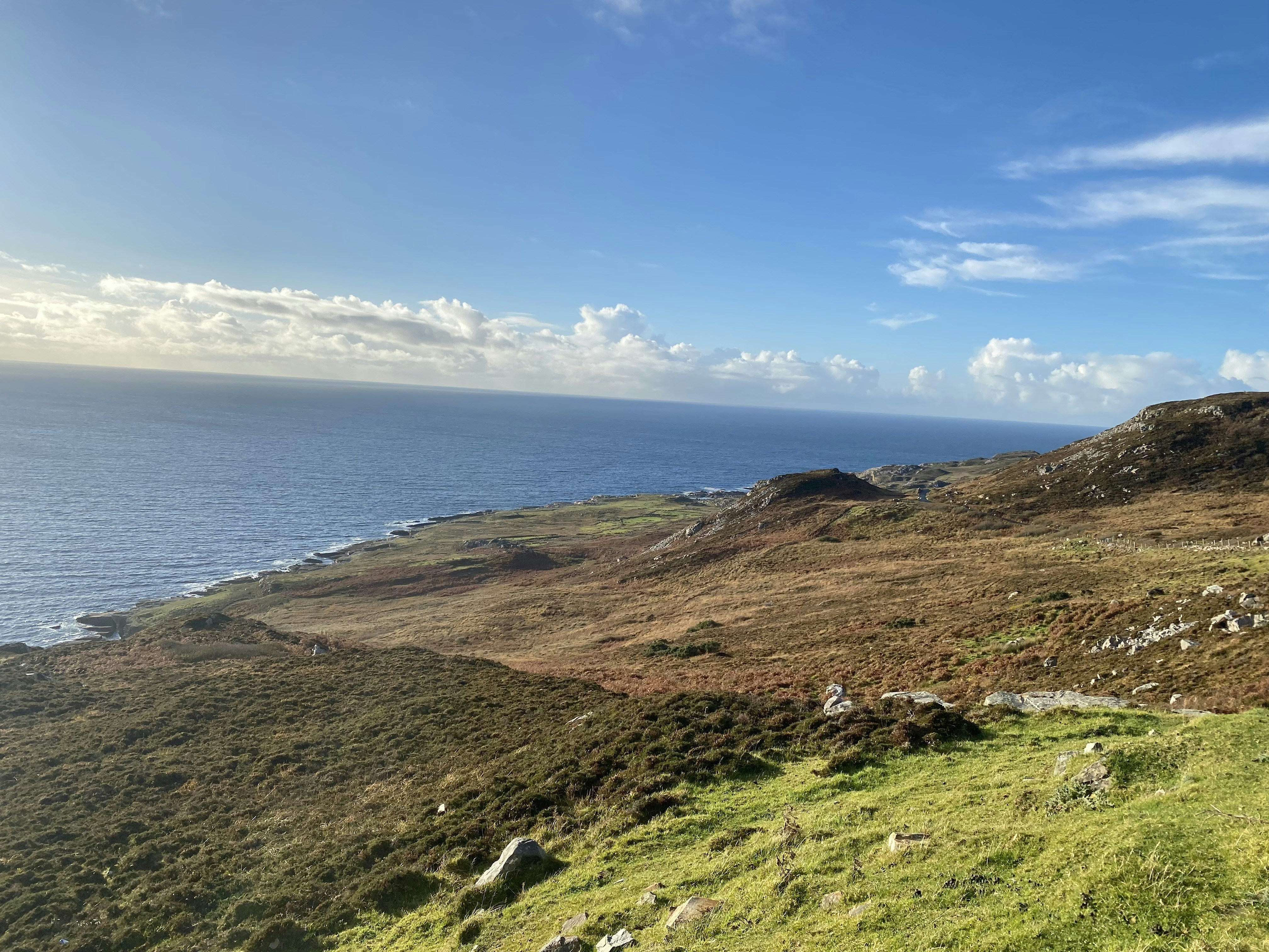 Vast coastal landscape with rolling hills leading to the ocean under a blue sky dotted with clouds.