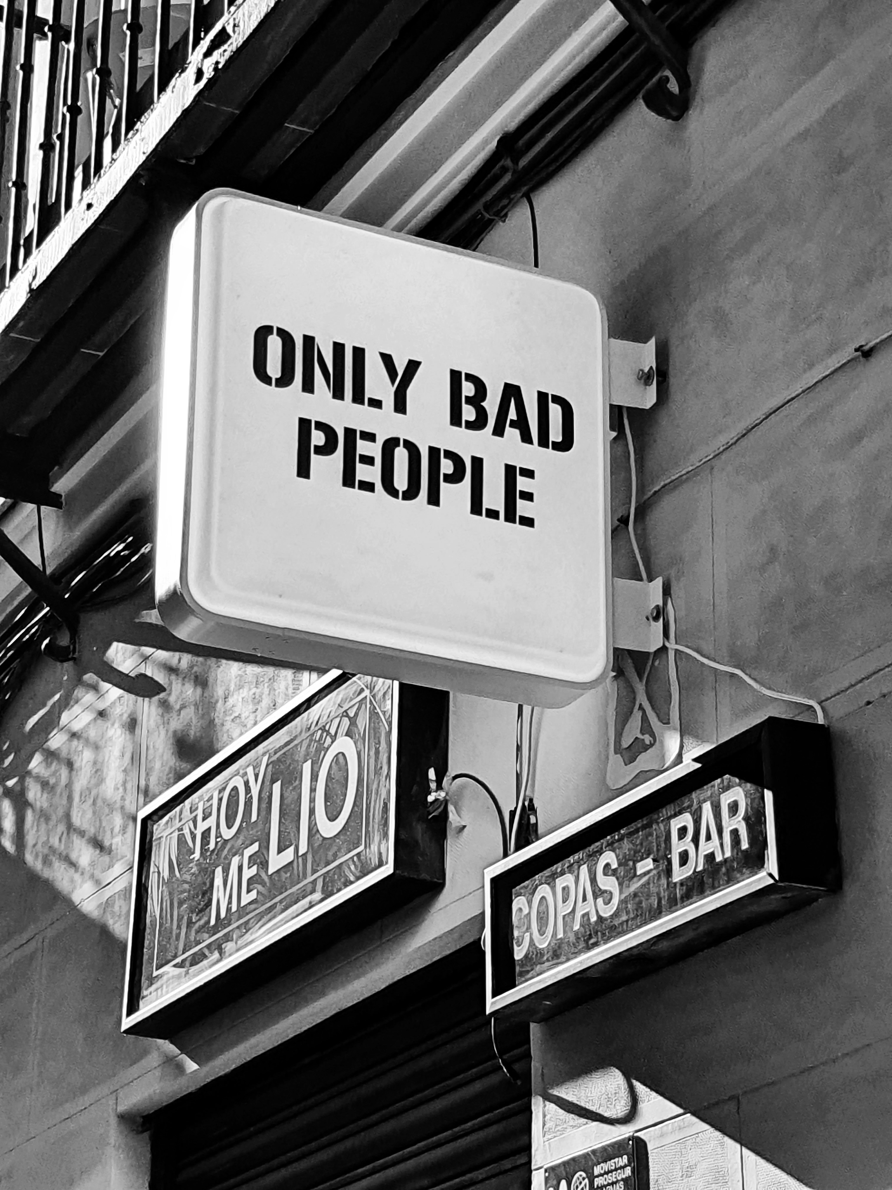 Illuminated sign reading 'ONLY BAD PEOPLE' juxtaposed with smaller signs in a monochrome setting, hinting at a lively bar atmosphere.