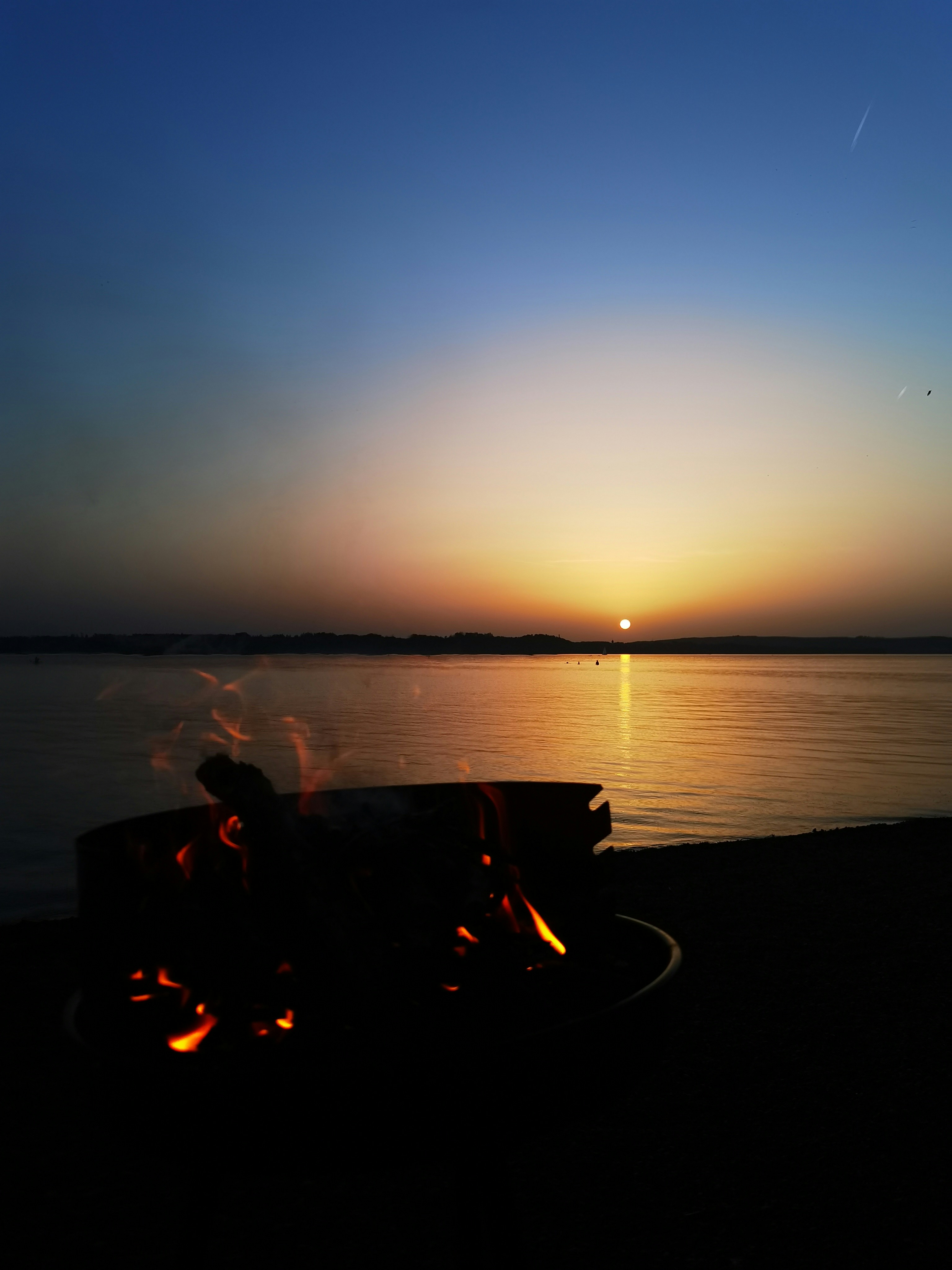 Flickering embers from a fire pit contrast against a tranquil sunset over a calm lake, with the sun dipping below the horizon.