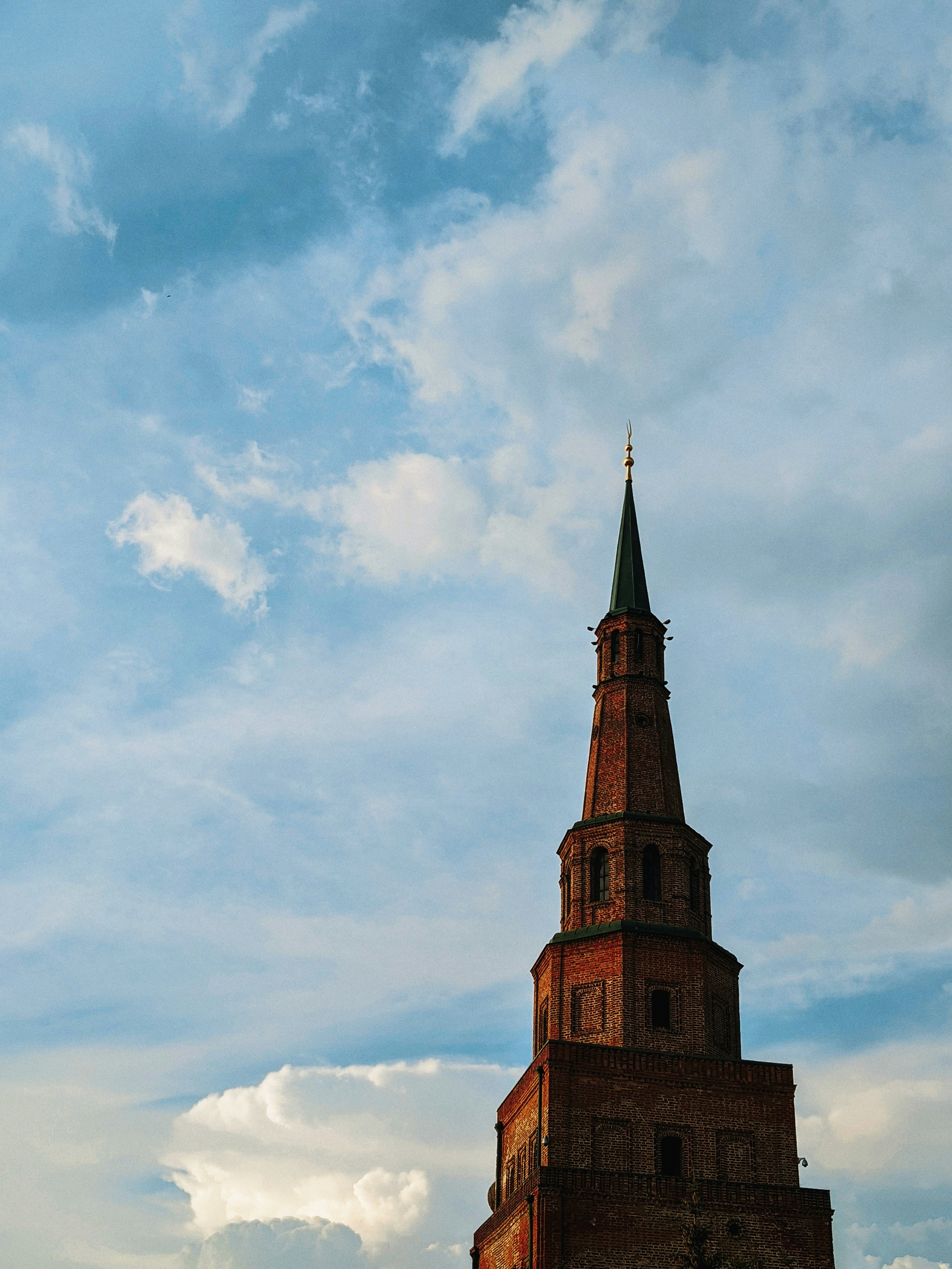 Historic brick tower with a green spire rises against a backdrop of blue sky and fluffy clouds.