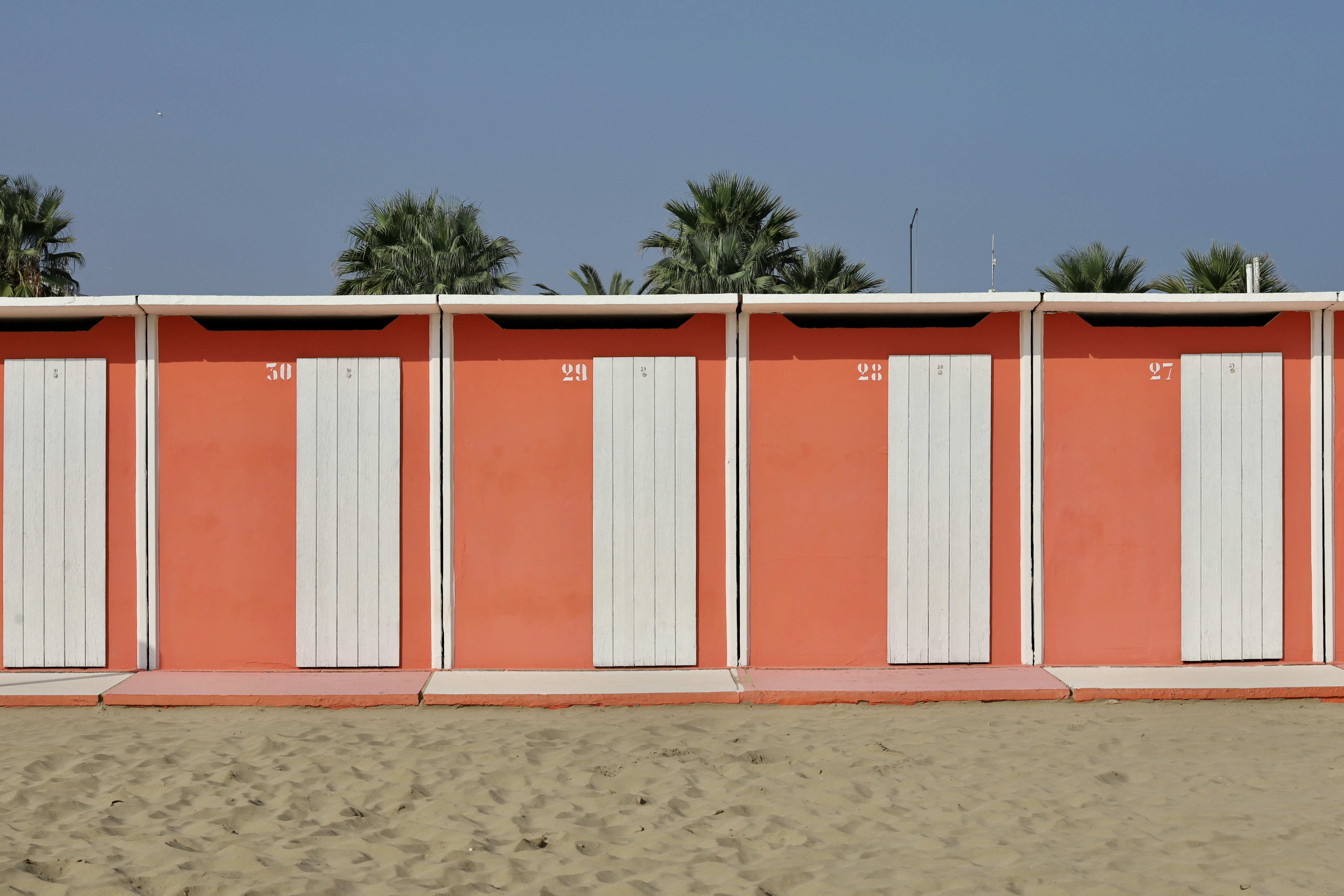 Bright orange beach huts with white doors line a sandy shore, framed by palm trees under a clear blue sky.