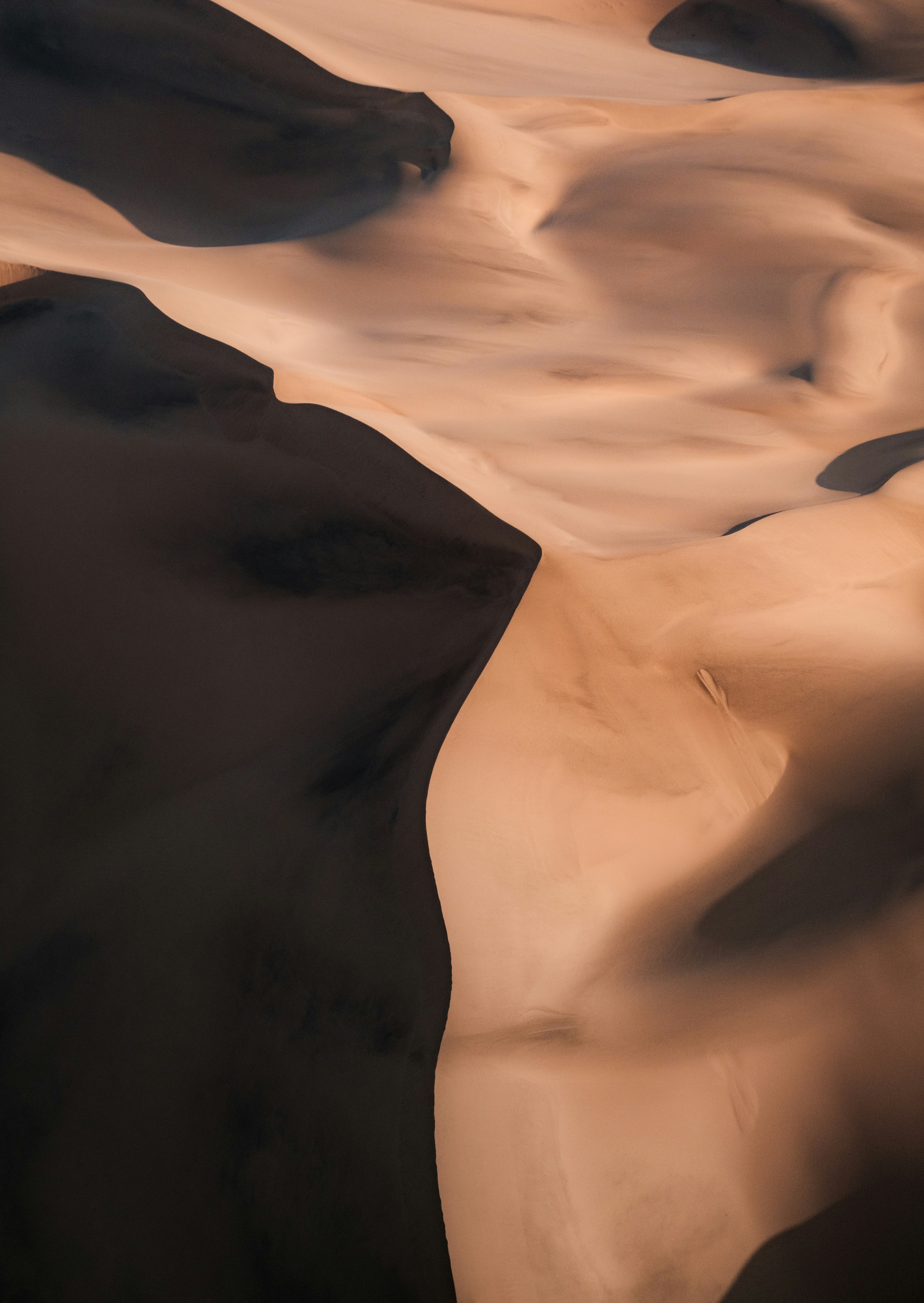 Wind-sculpted dunes glow in warm peach tones with dramatic shadowed valleys, creating an abstract desert composition. Landscape photograph highlighting sculptural curves and negative space.