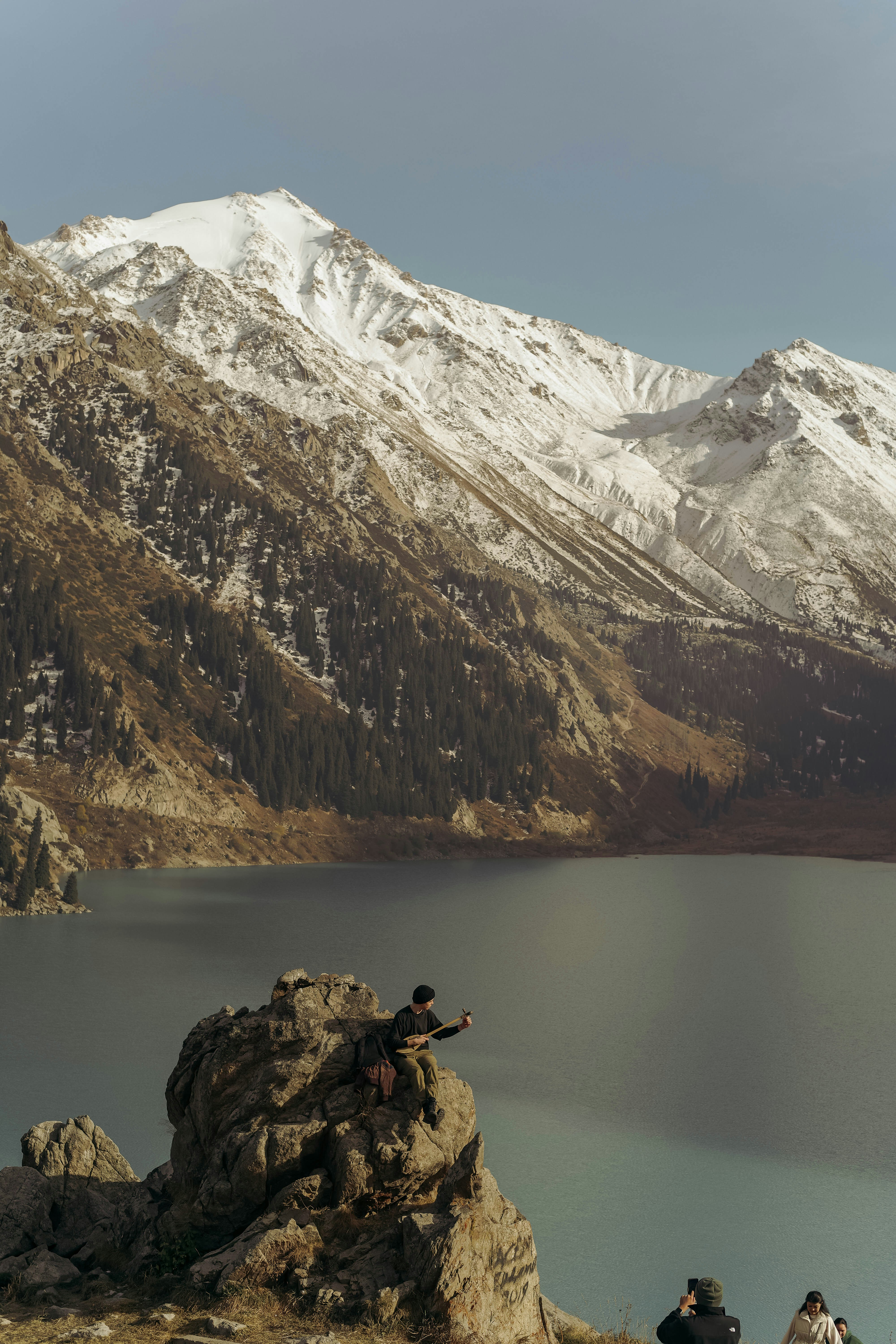 Alpine Sentinel on the Rock: A lone climber gazing over a glacial lake beneath snow-dusted peaks.
