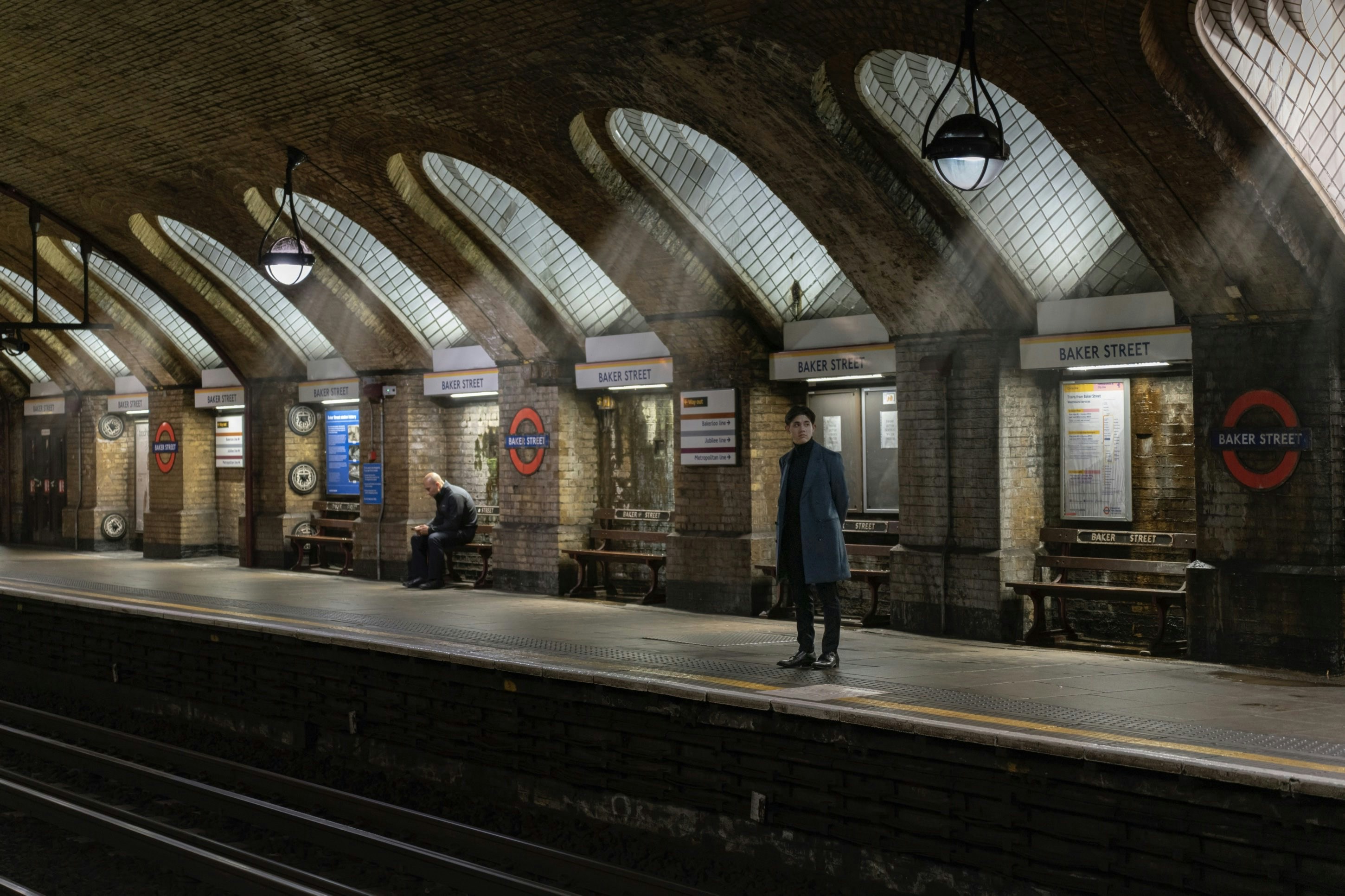 A lone figure stands on the platform of Baker Street station, illuminated by soft beams of light filtering through the arched windows, while a seated passenger waits in the background.