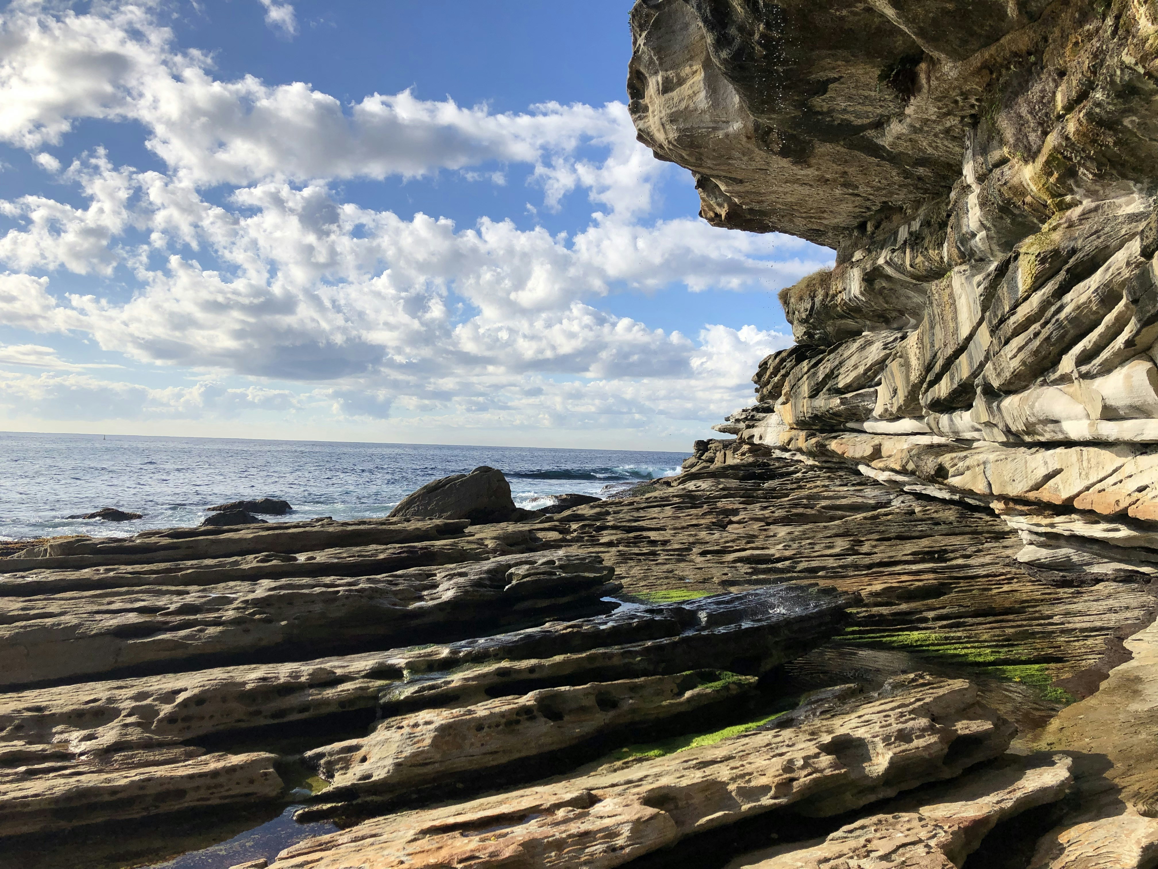 Intricate rock formations along a coastal shoreline with gentle waves lapping against the stones under a partly cloudy sky.