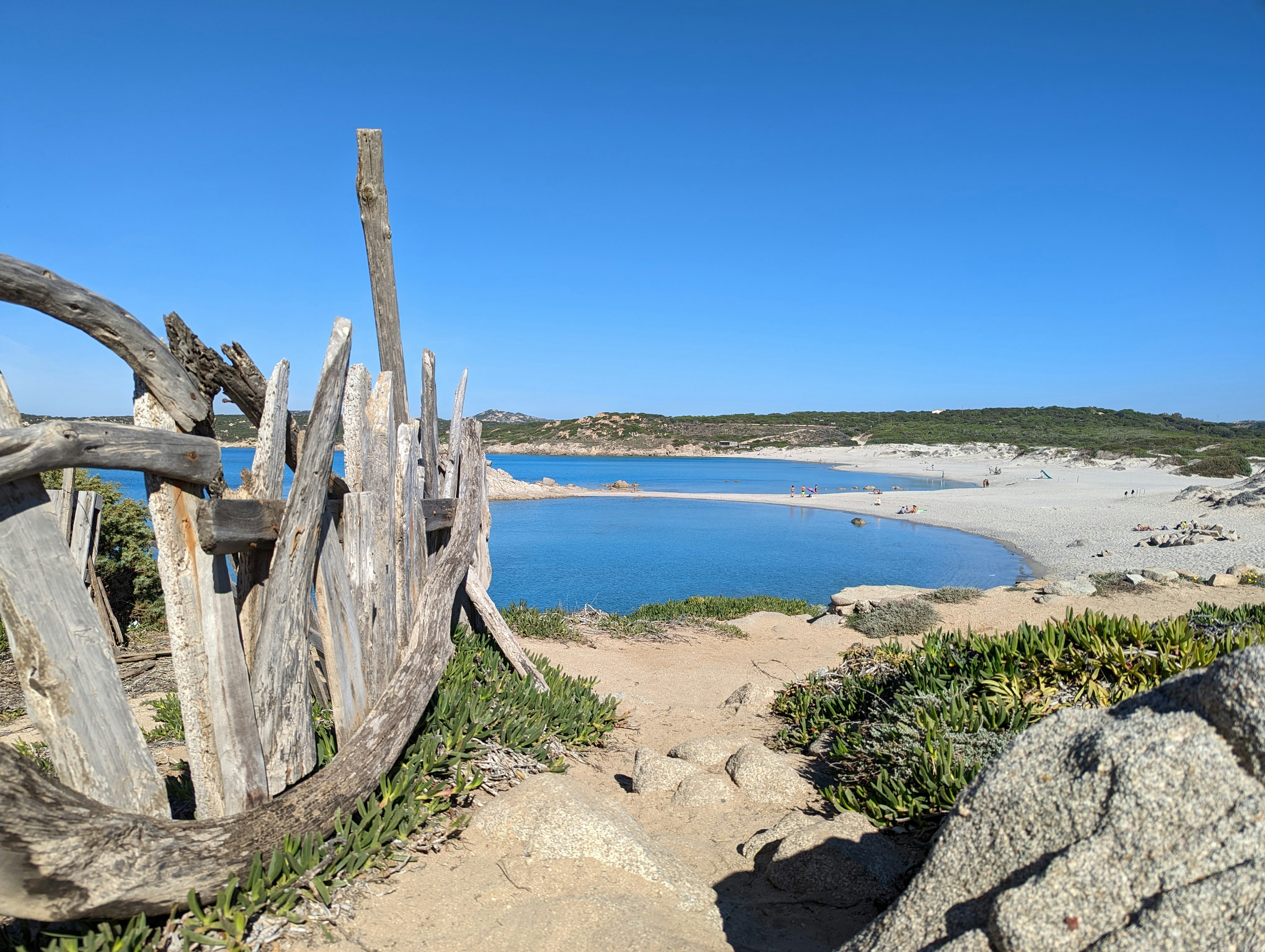 A rustic wooden fence frames a tranquil beach scene with clear blue waters and gentle sandy shores under a bright blue sky.