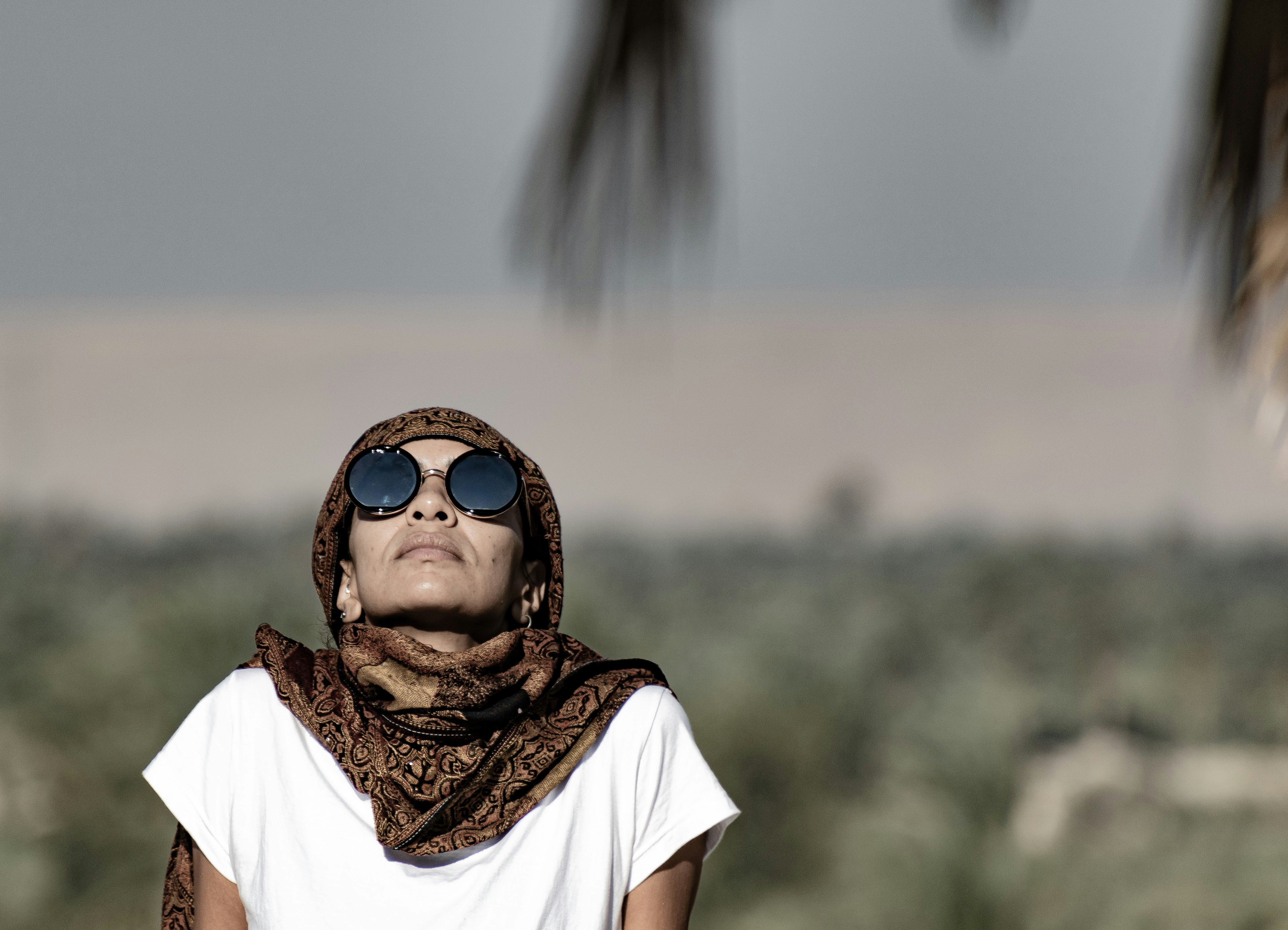 A woman wearing sunglasses and a patterned scarf gazes upward, surrounded by a lush landscape. The scene conveys a sense of tranquility and connection to nature.