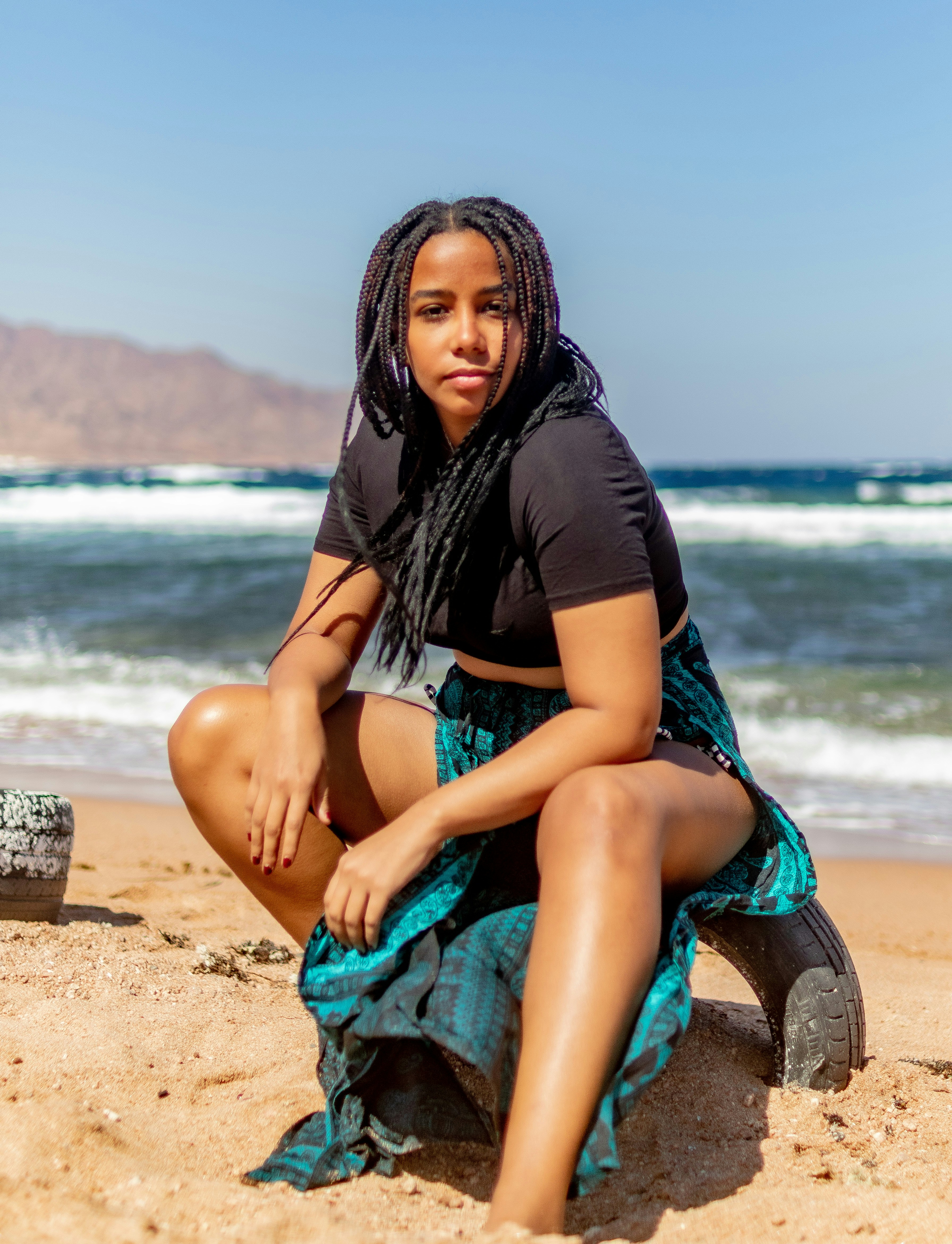 Person sitting on a sandy beach with waves crashing in the background under a clear blue sky.