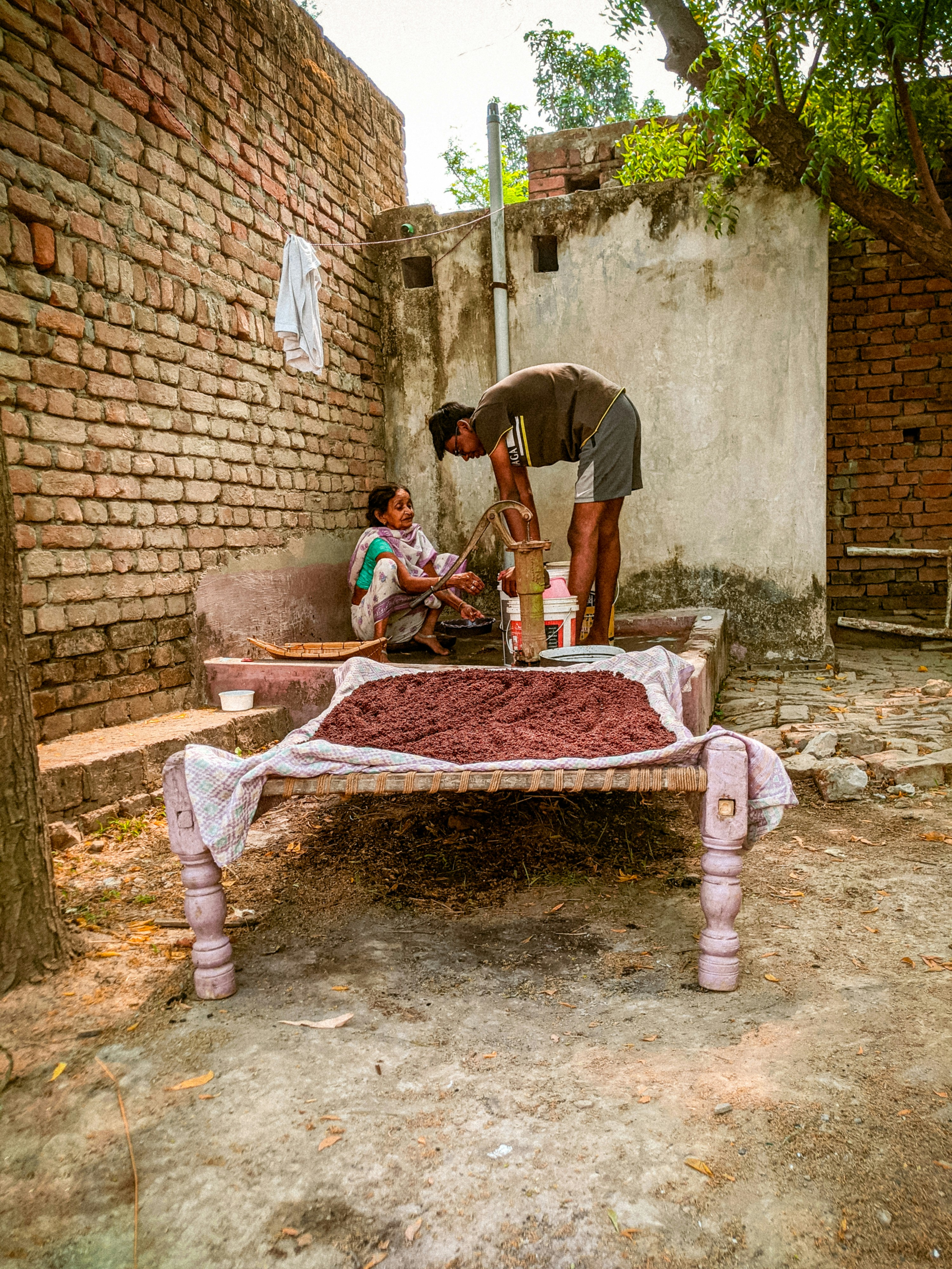 A man and woman engaged in a traditional preparation process, surrounded by rustic brick walls and natural elements. The scene captures the essence of cultural practices in a rural setting.