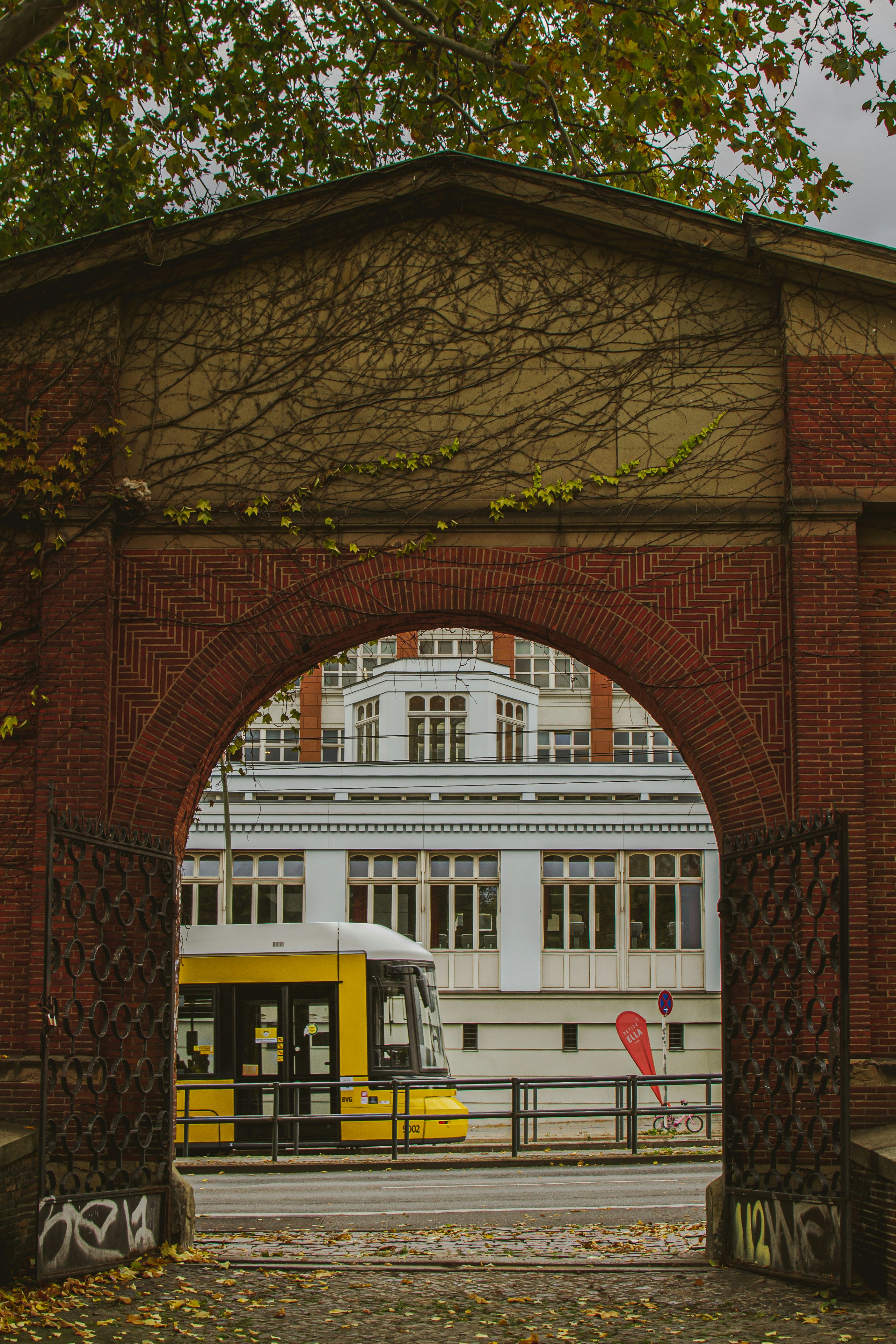 Yellow tram passing through an archway, framed by ivy-covered brickwork and modern architecture in the background.