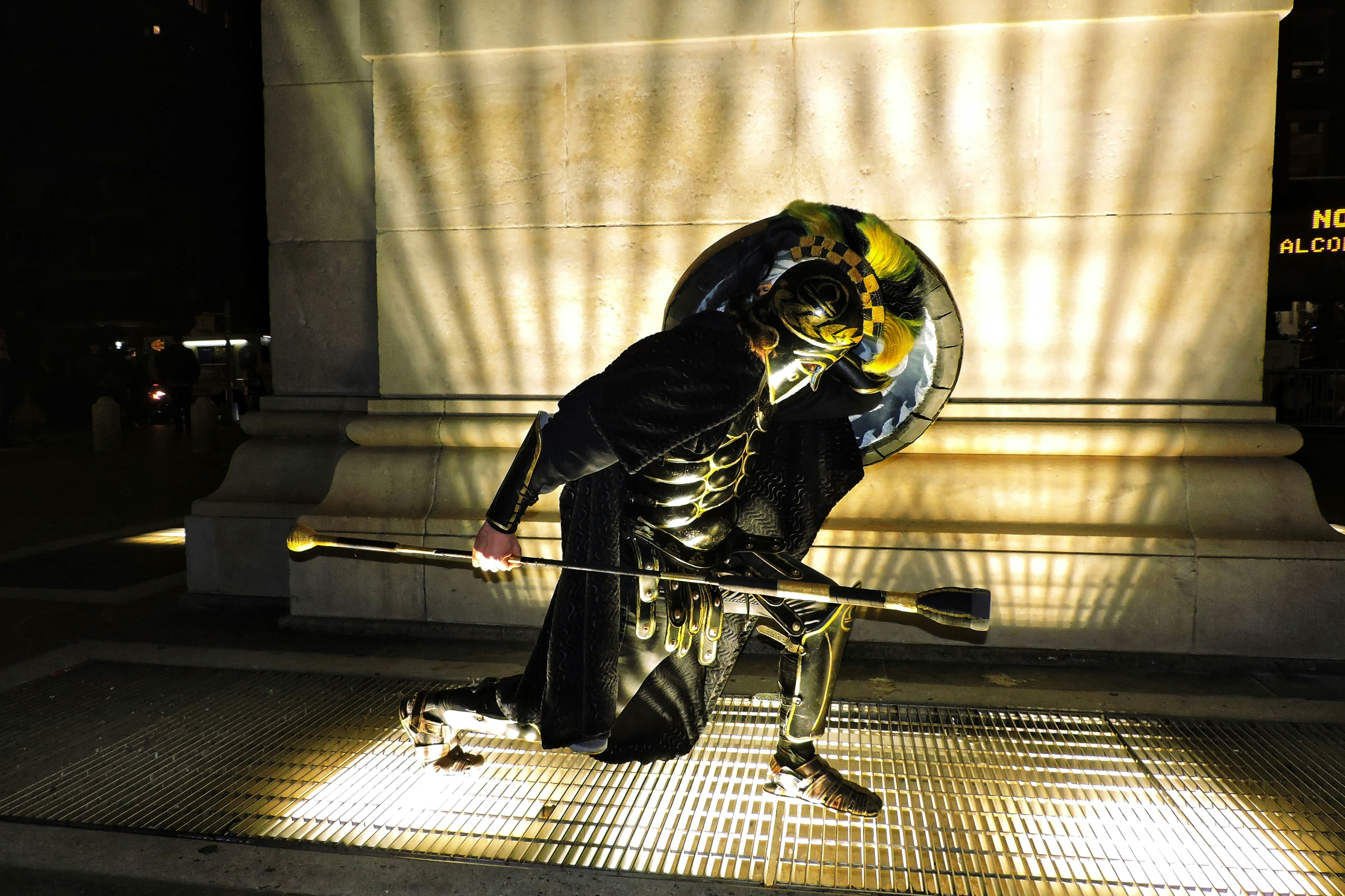 A dynamic street performer in a warrior costume, poised with a spear, illuminated by dramatic lighting against a stone backdrop.