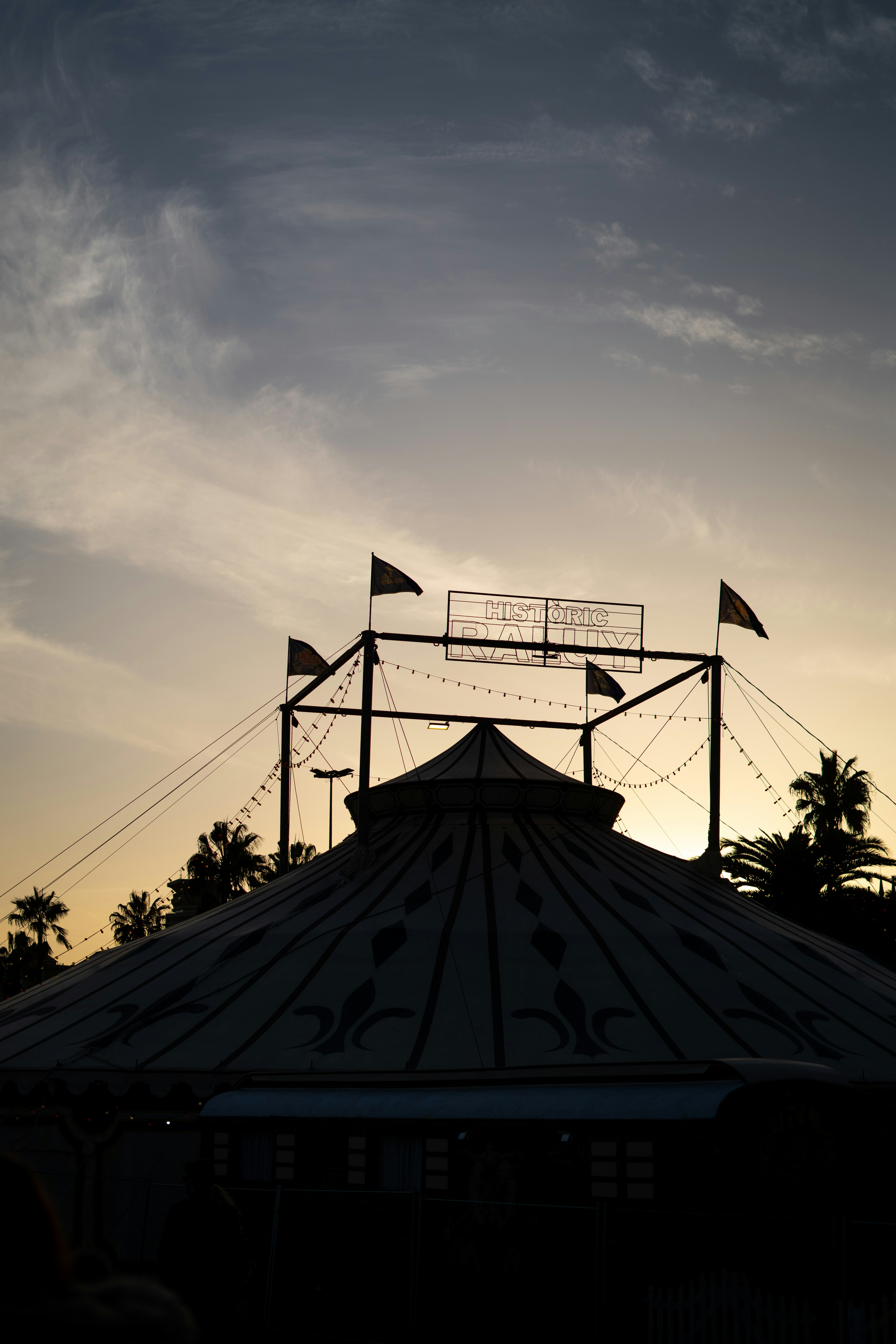 Historic carnival tent silhouetted against a colorful sunset, with flags fluttering in the breeze. The scene evokes nostalgia and excitement.