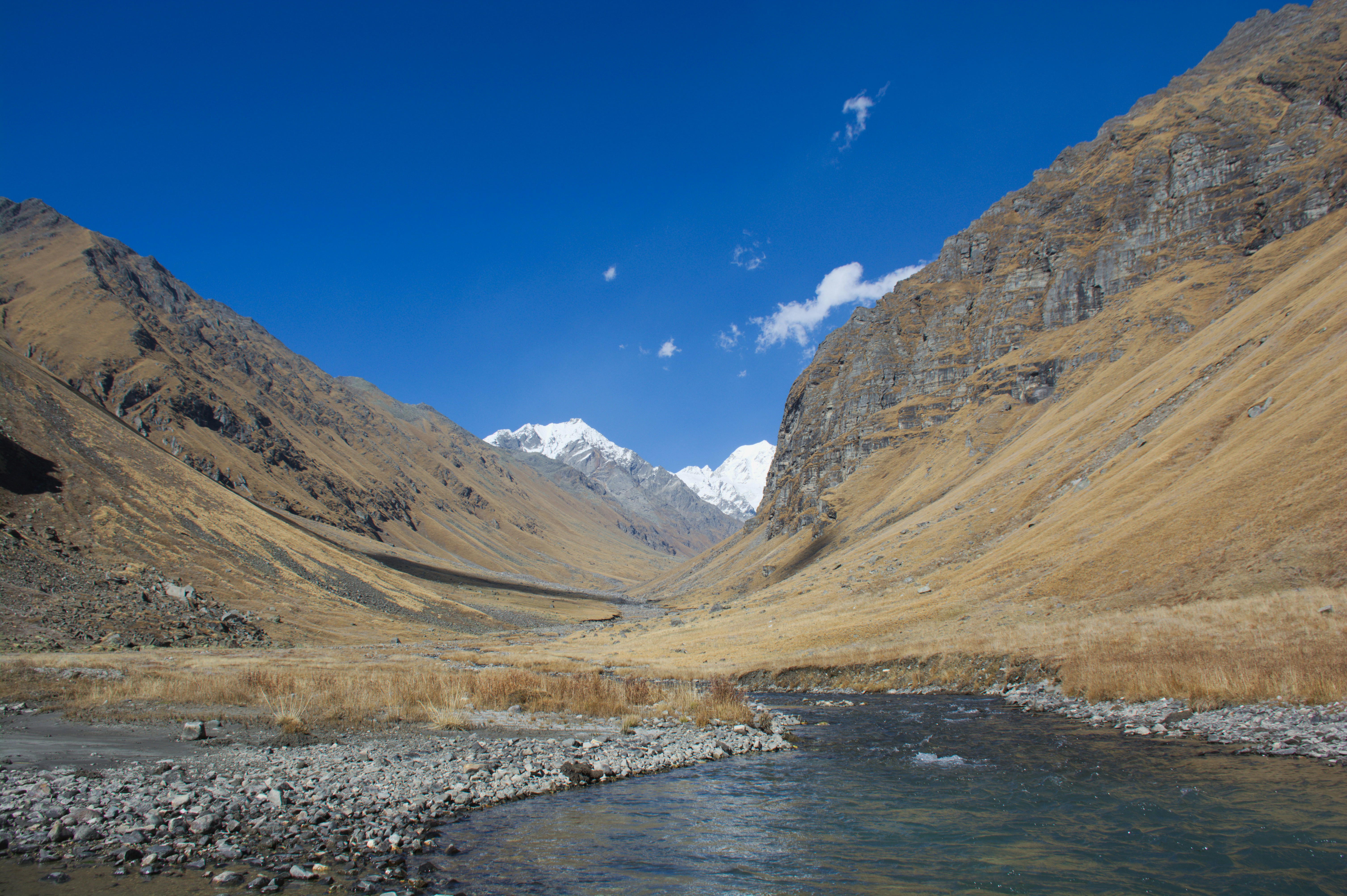 Mountain valley view with a river flowing under a clear blue sky, leading towards snow-capped peaks.