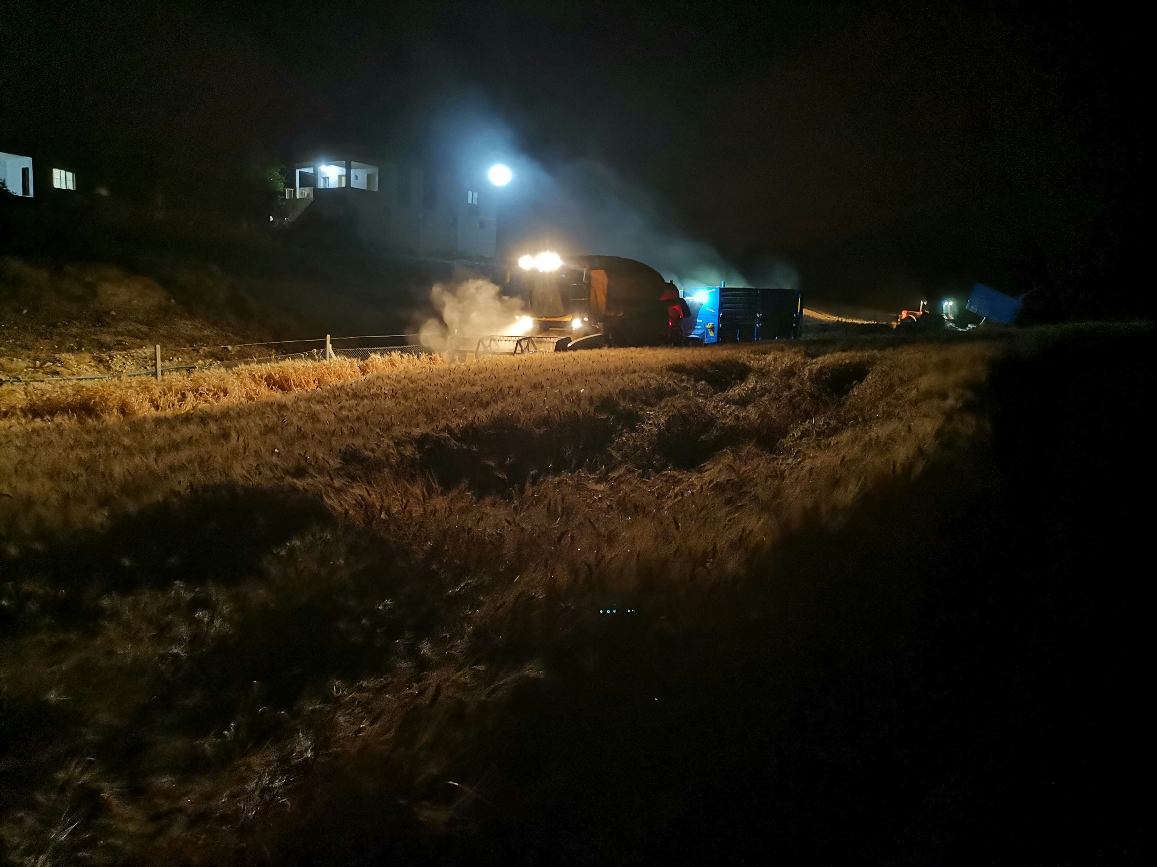 A tractor working under artificial lights on a field at night, surrounded by wisps of steam and distant buildings. The scene captures the essence of agricultural labor after dark.