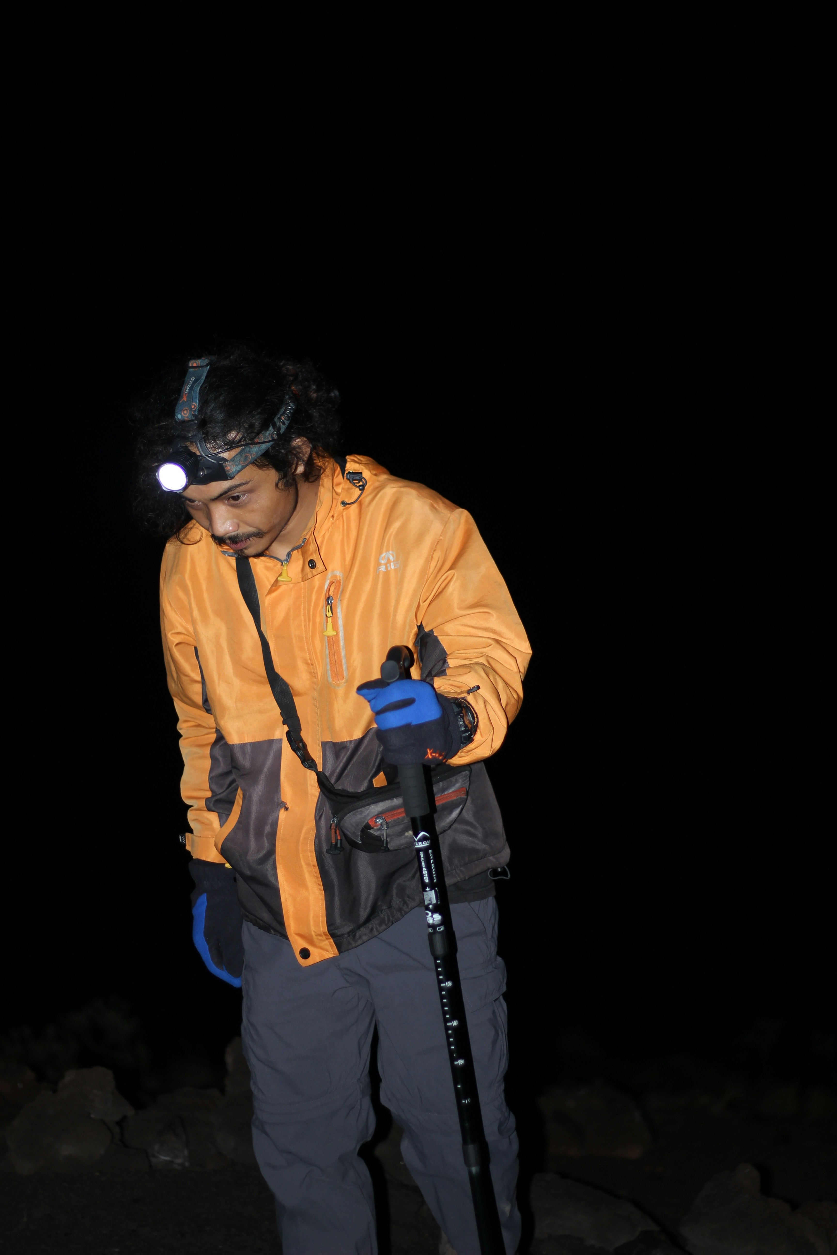 Hiker in a bright orange jacket and blue gloves, focused on the ground while using a trekking pole under a headlamp's glow during nighttime exploration.