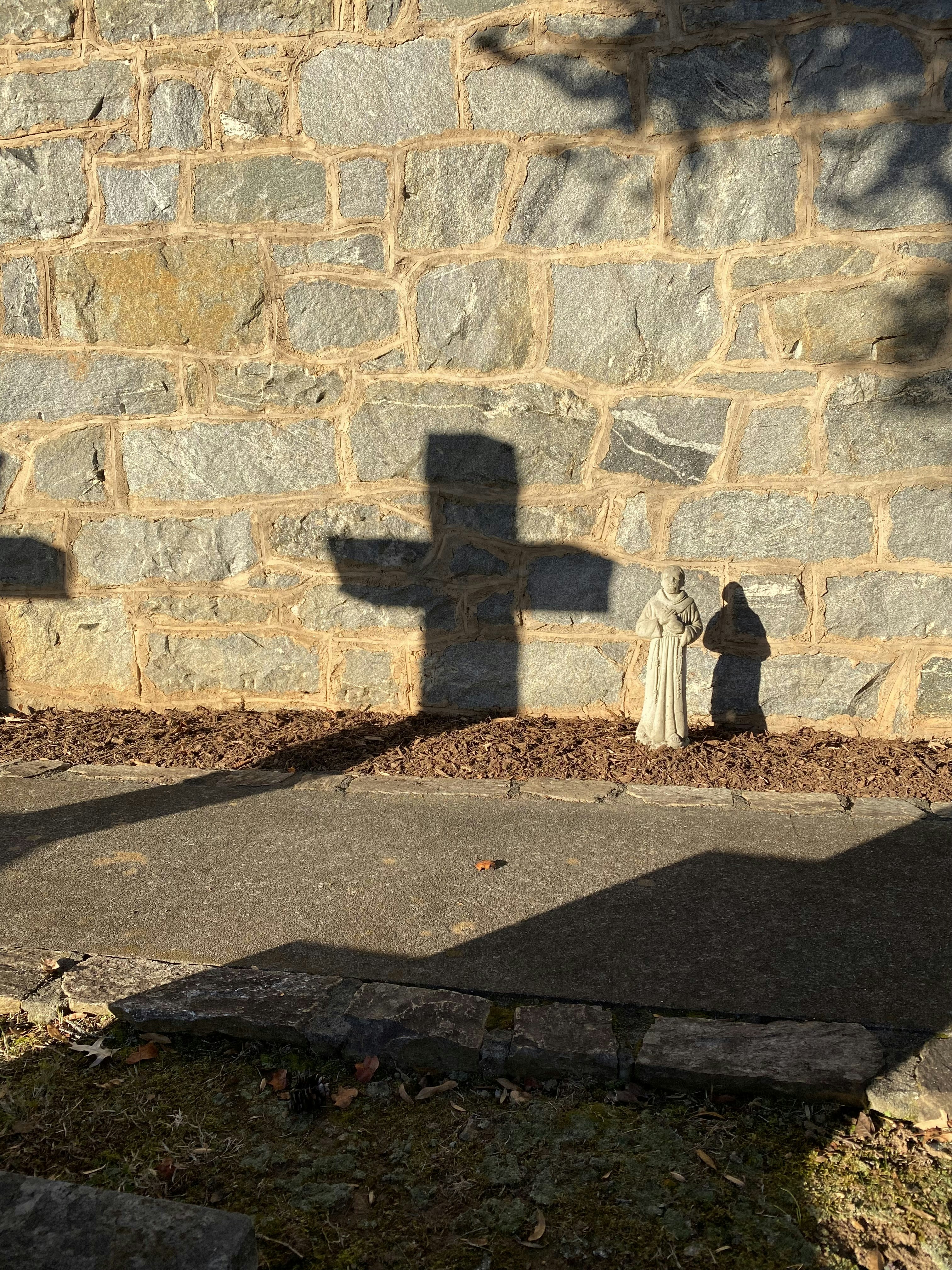 A stone wall casts dramatic shadows of crosses alongside a small statue, highlighting themes of faith and memory.