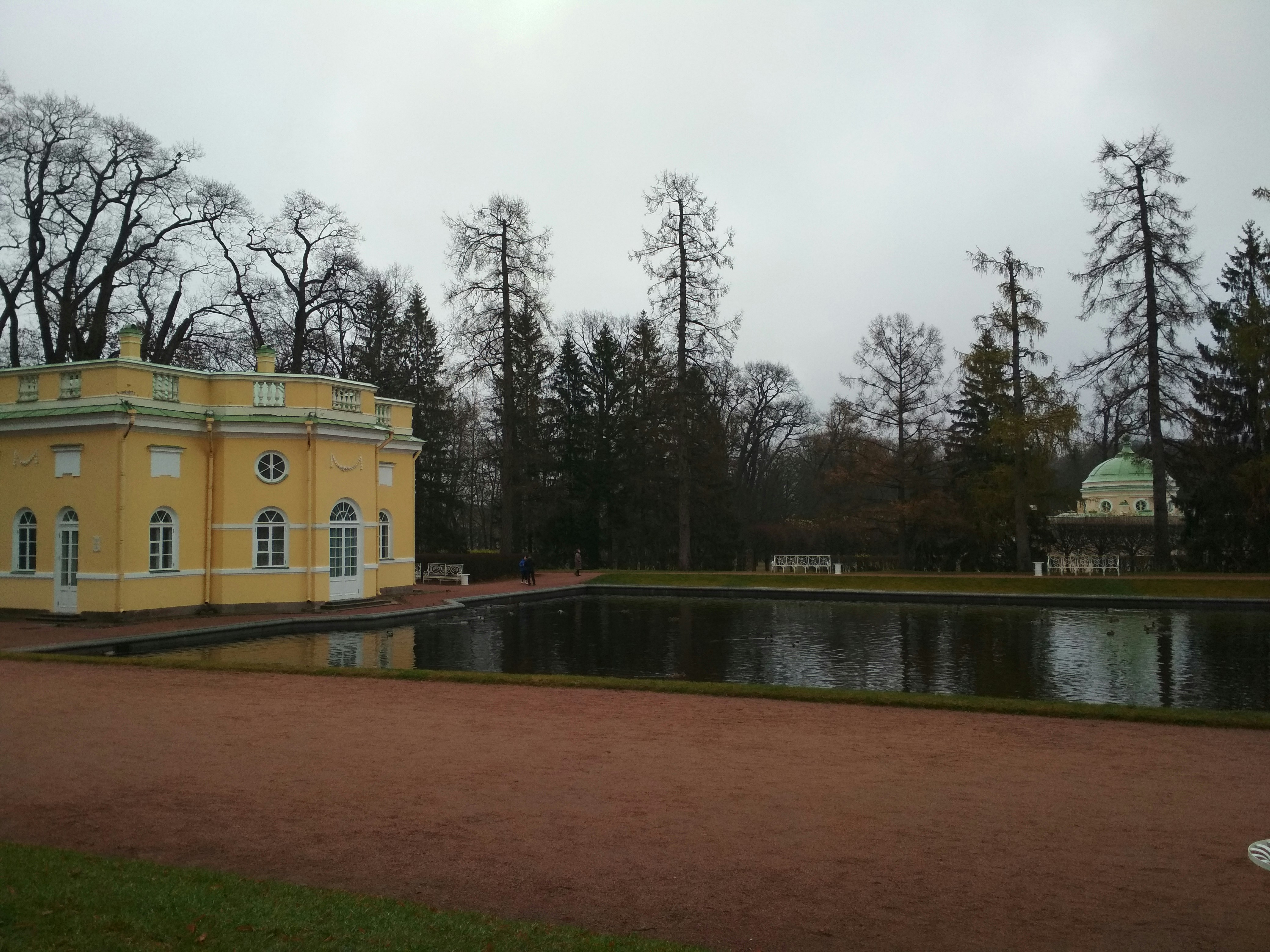 Yellow bathhouse beside a serene pond surrounded by tall, bare trees under an overcast sky.