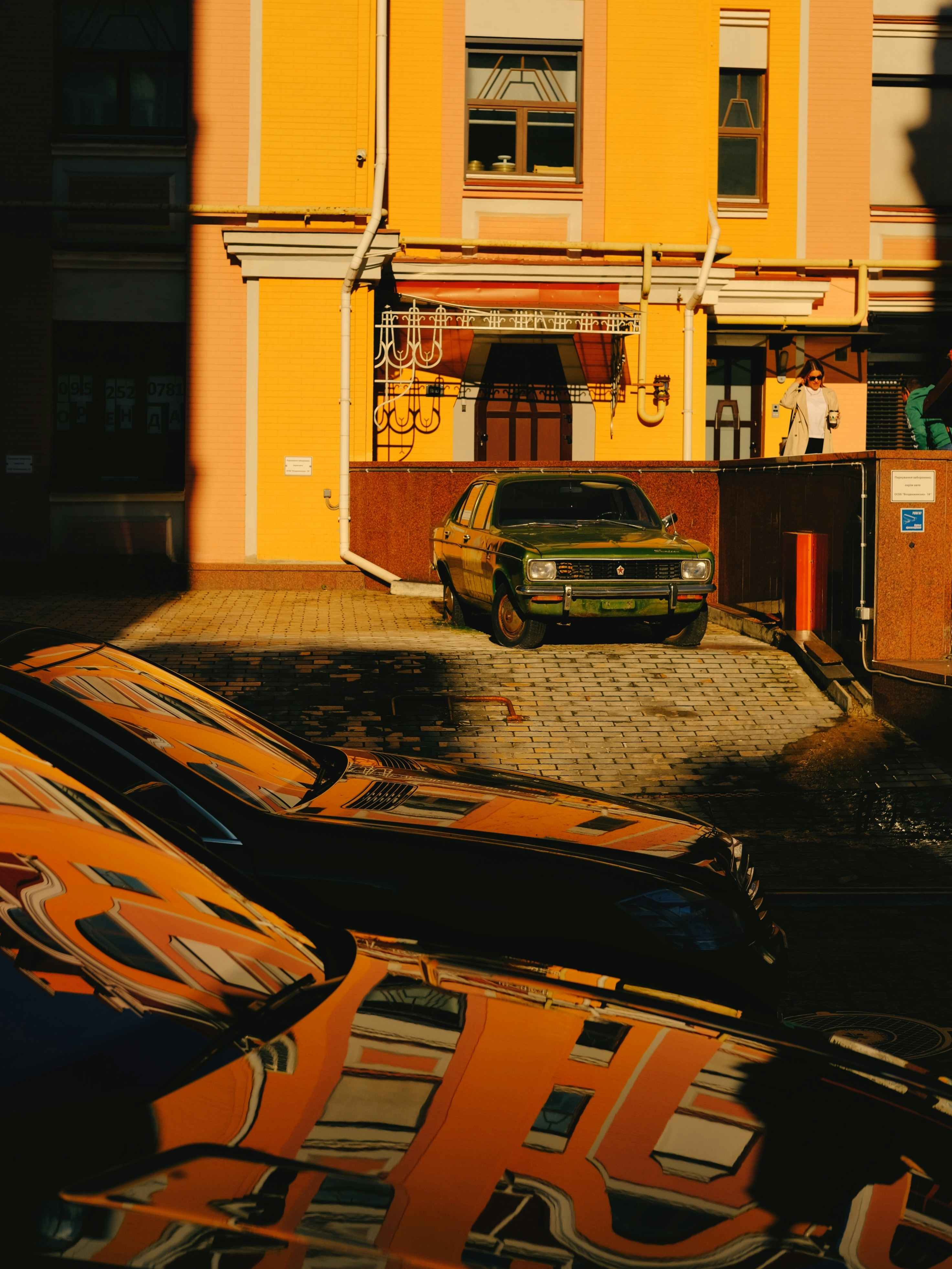 A classic green car parked in a sunlit courtyard, reflecting vibrant colors from the surrounding architecture. The scene captures a blend of nostalgia and contemporary urban life.