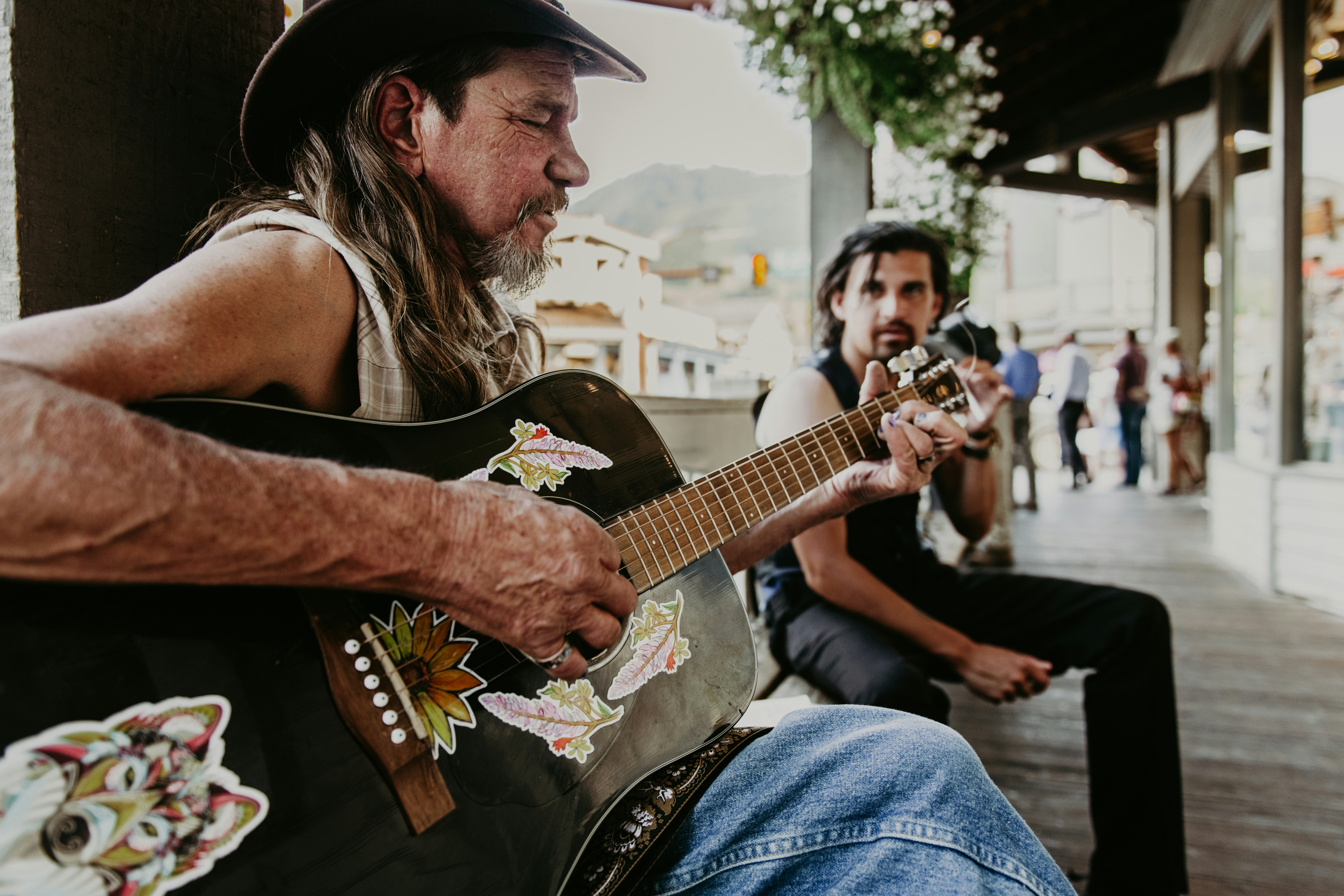 Street musician with a sticker-adorned guitar performs on a bustling city sidewalk, another person listens nearby.